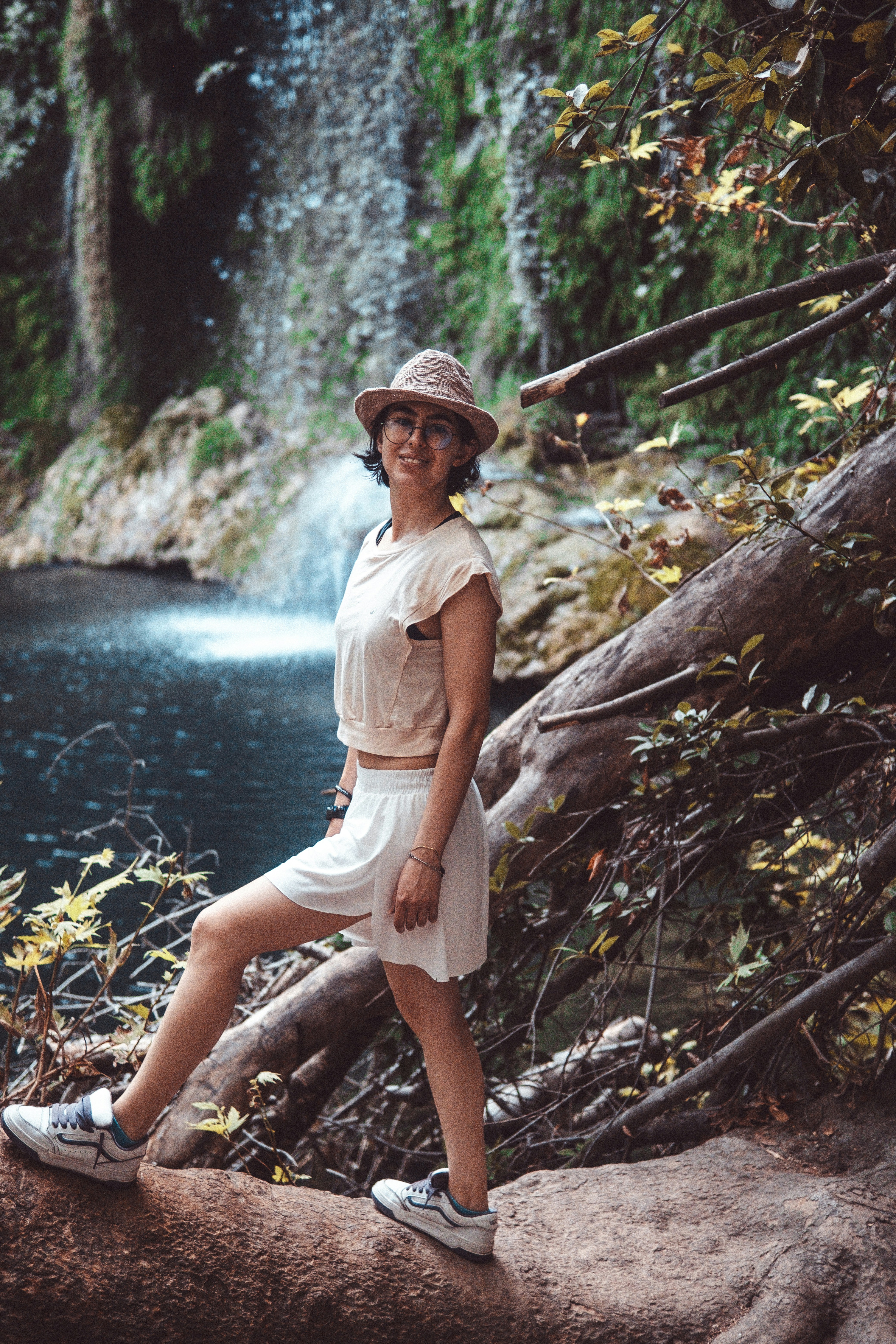 A woman in light clothing and a hat stands on a fallen log beside a tranquil waterfall, framed by lush greenery and rocky terrain.