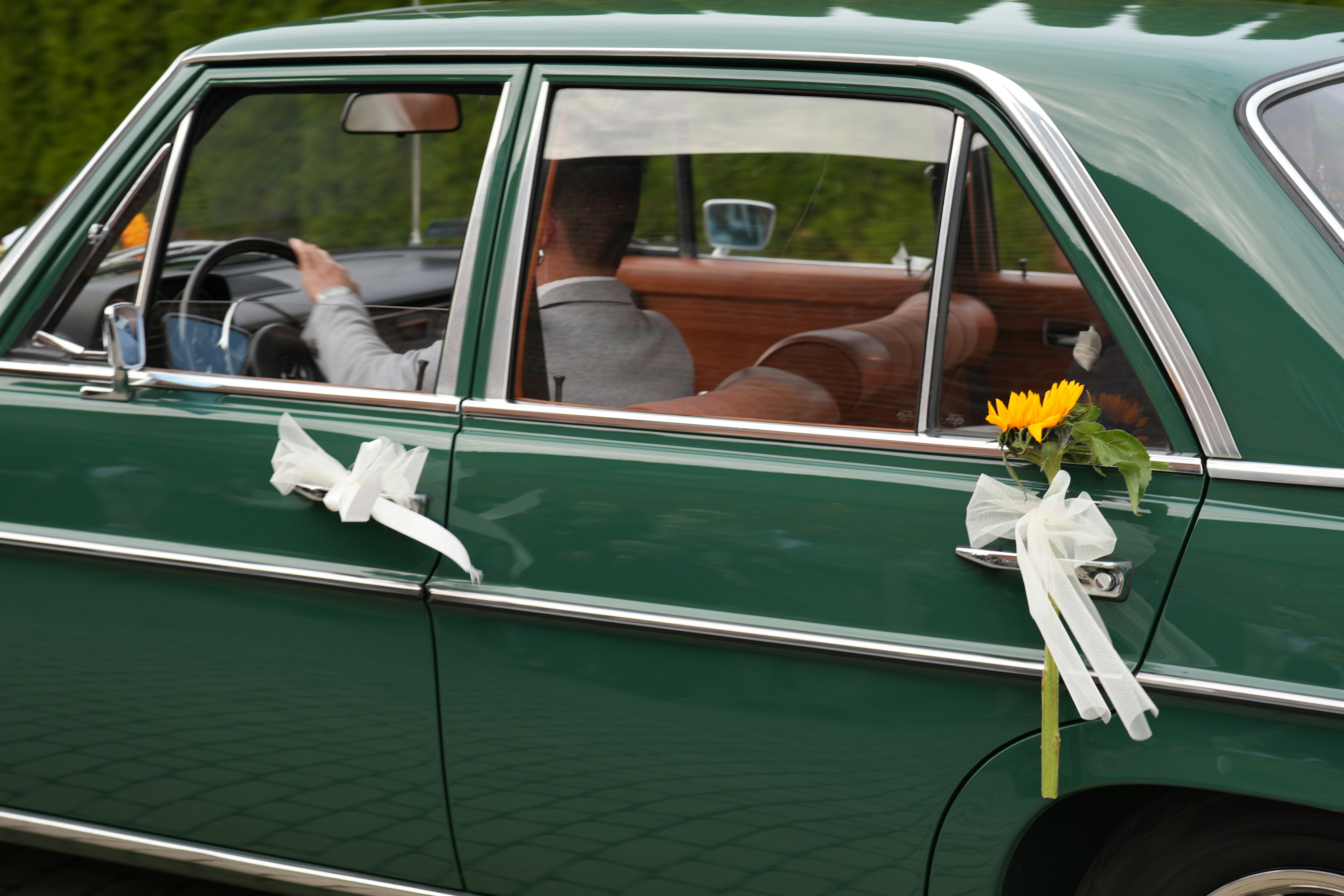 A vintage green car with white ribbons and a yellow flower adornment sits parked; the scene shows a couple inside, with the driver and a passenger glimpsed through the windows.