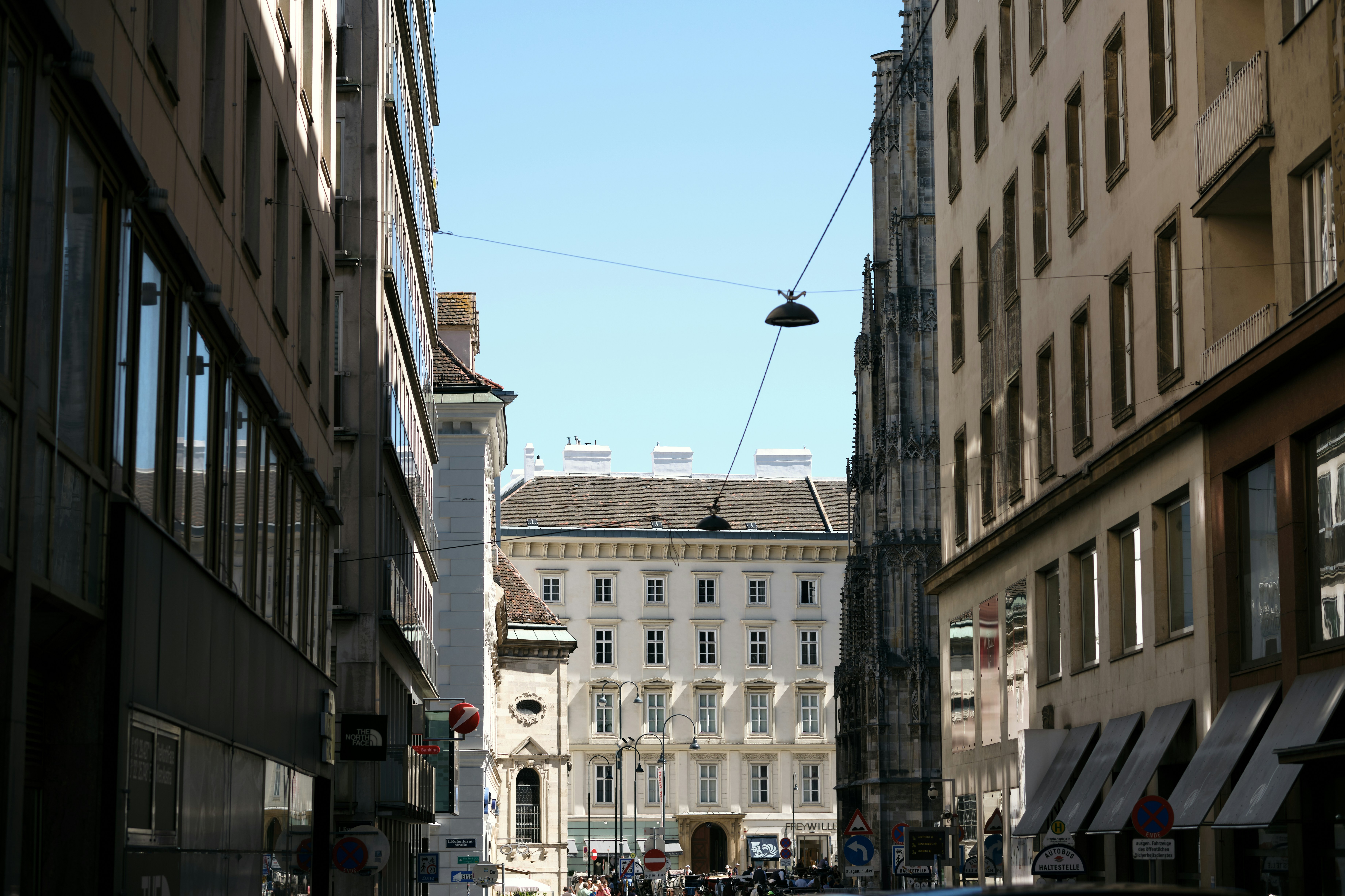 A sunlit street leads toward a grand city hall framed by tall buildings on either side, capturing the architectural depth and bustling urban scene.