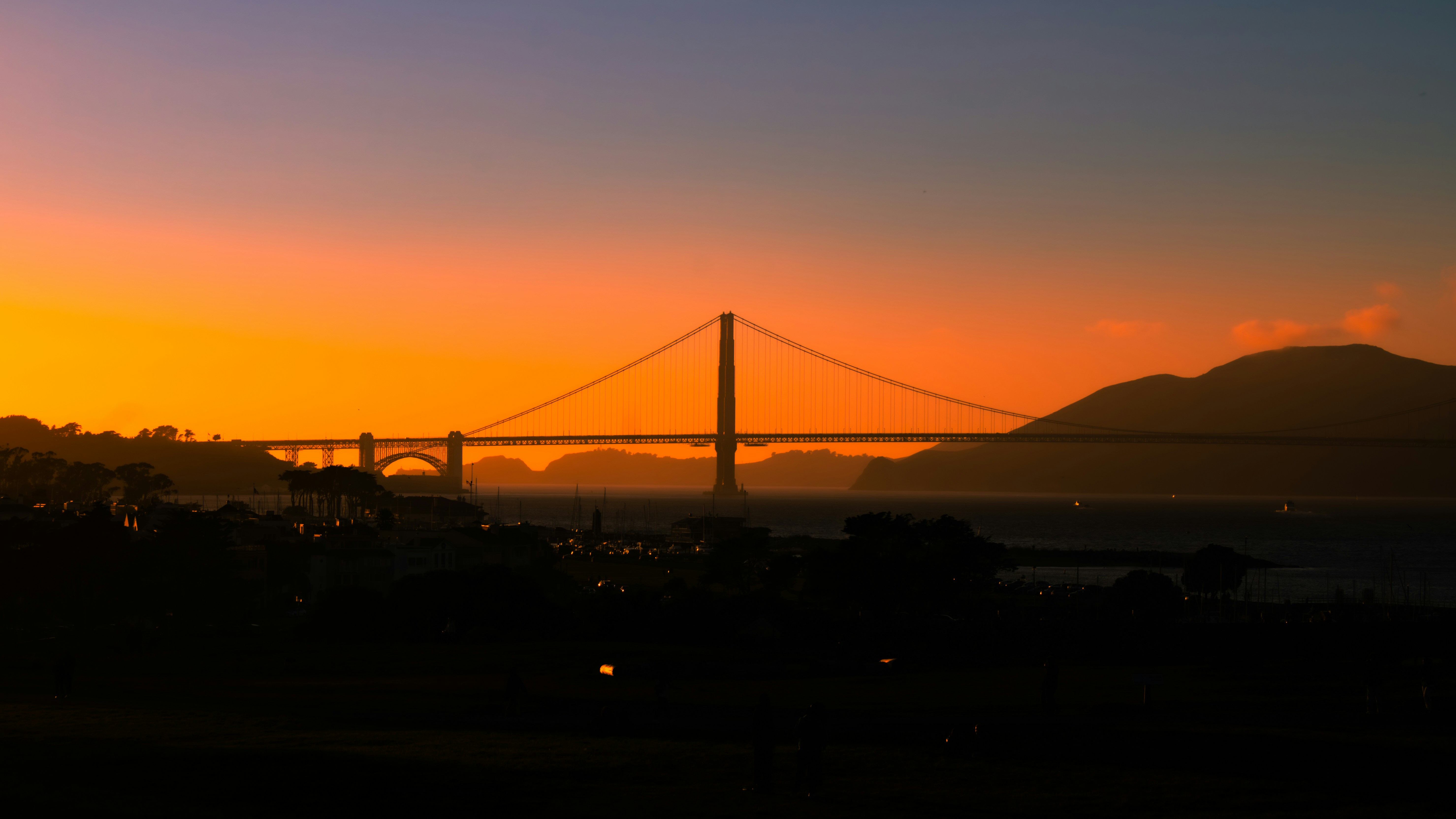 Silhouetted bridge spans a glowing horizon at sunset, with distant hills and a calm water foreground.