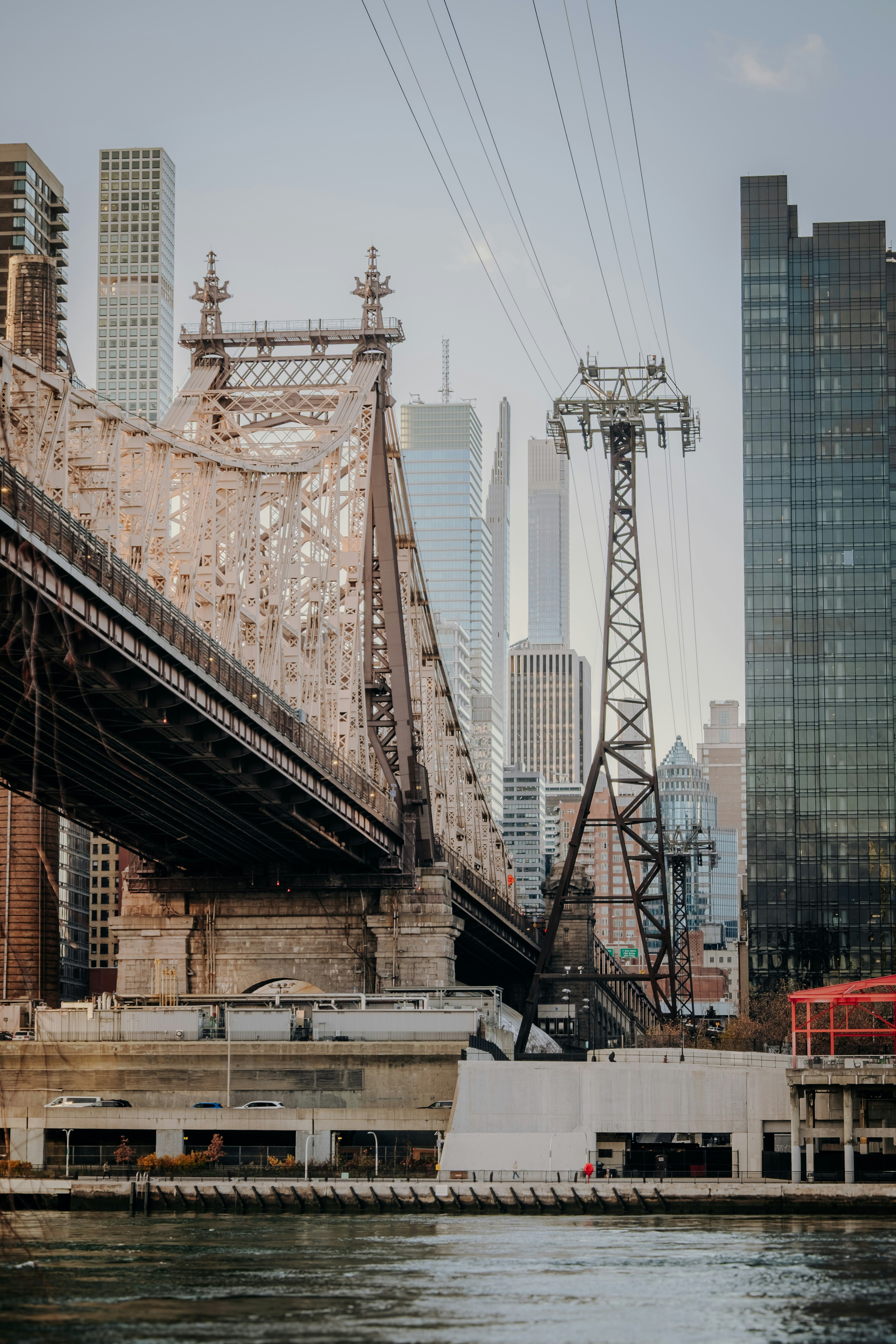 Steel bridge spans the foreground with a river beneath and a skyline of modern towers in the background, capturing city life from the waterfront. This is a photographic composition of urban architecture.