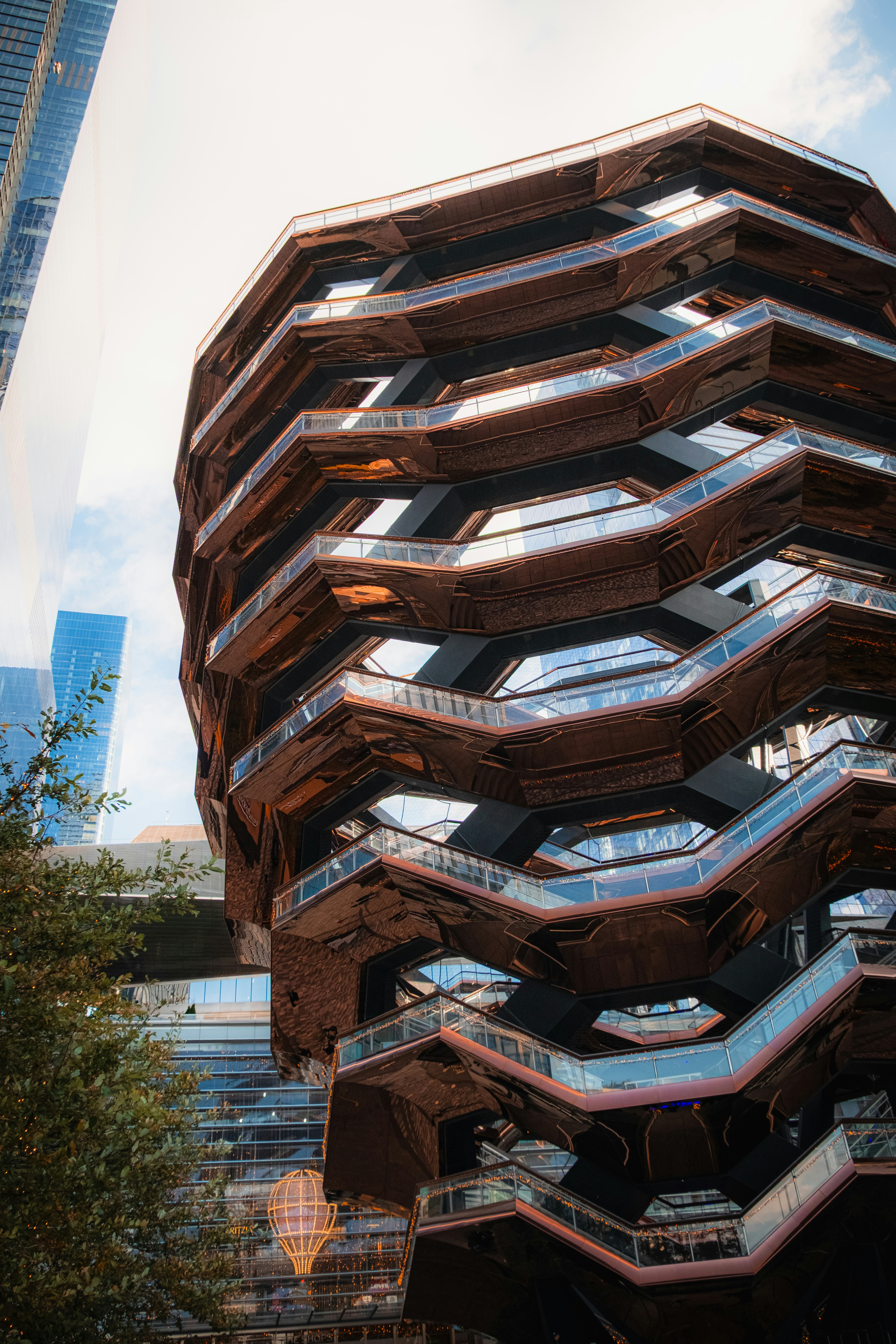 A towering, copper-toned architectural structure with stacked angular balconies climbs upward, reflecting light and surrounding skyline.