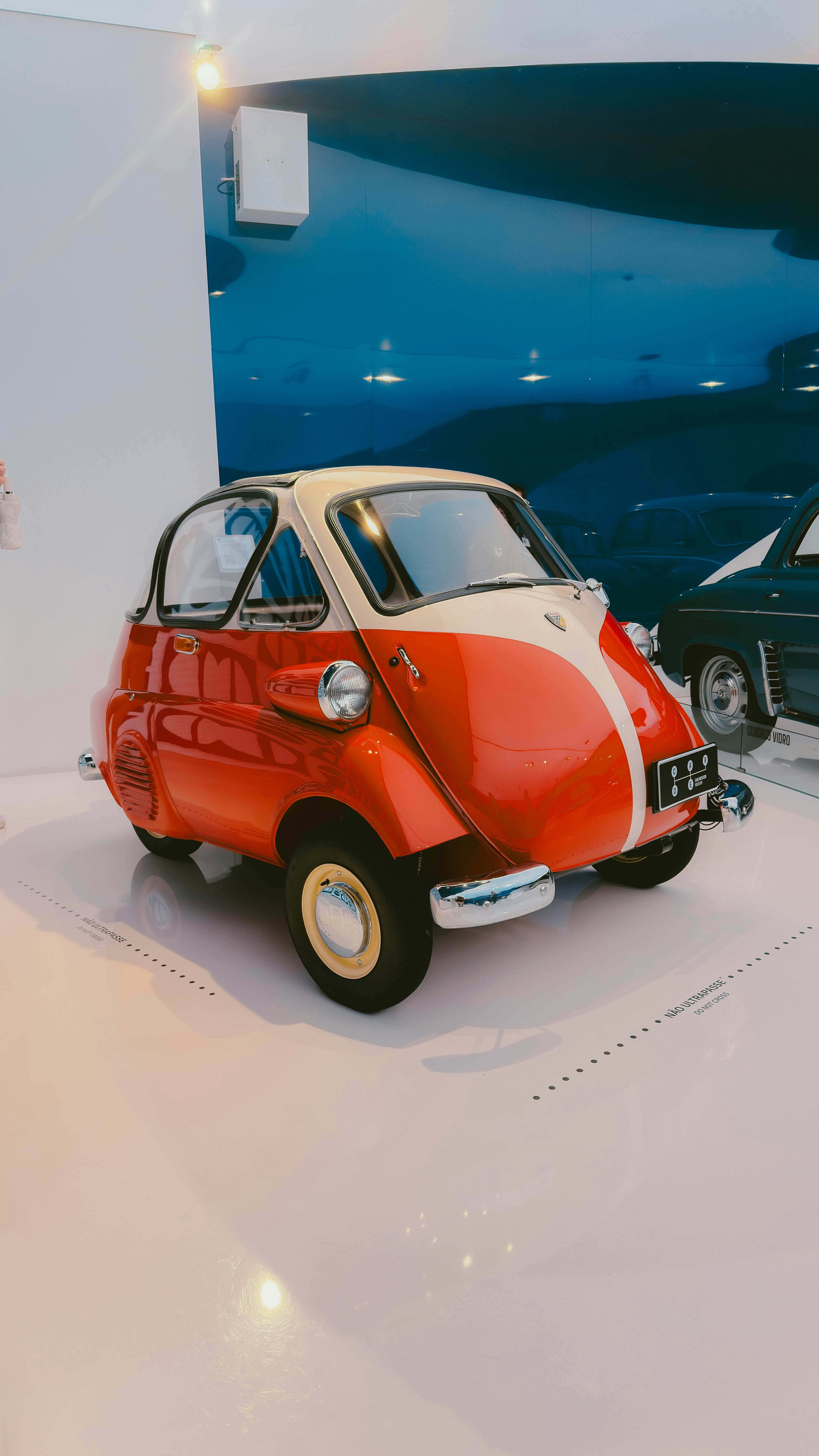 A vintage red microcar with white racing stripe on a glossy white pedestal in a showroom, framed by a blue reflective backdrop.