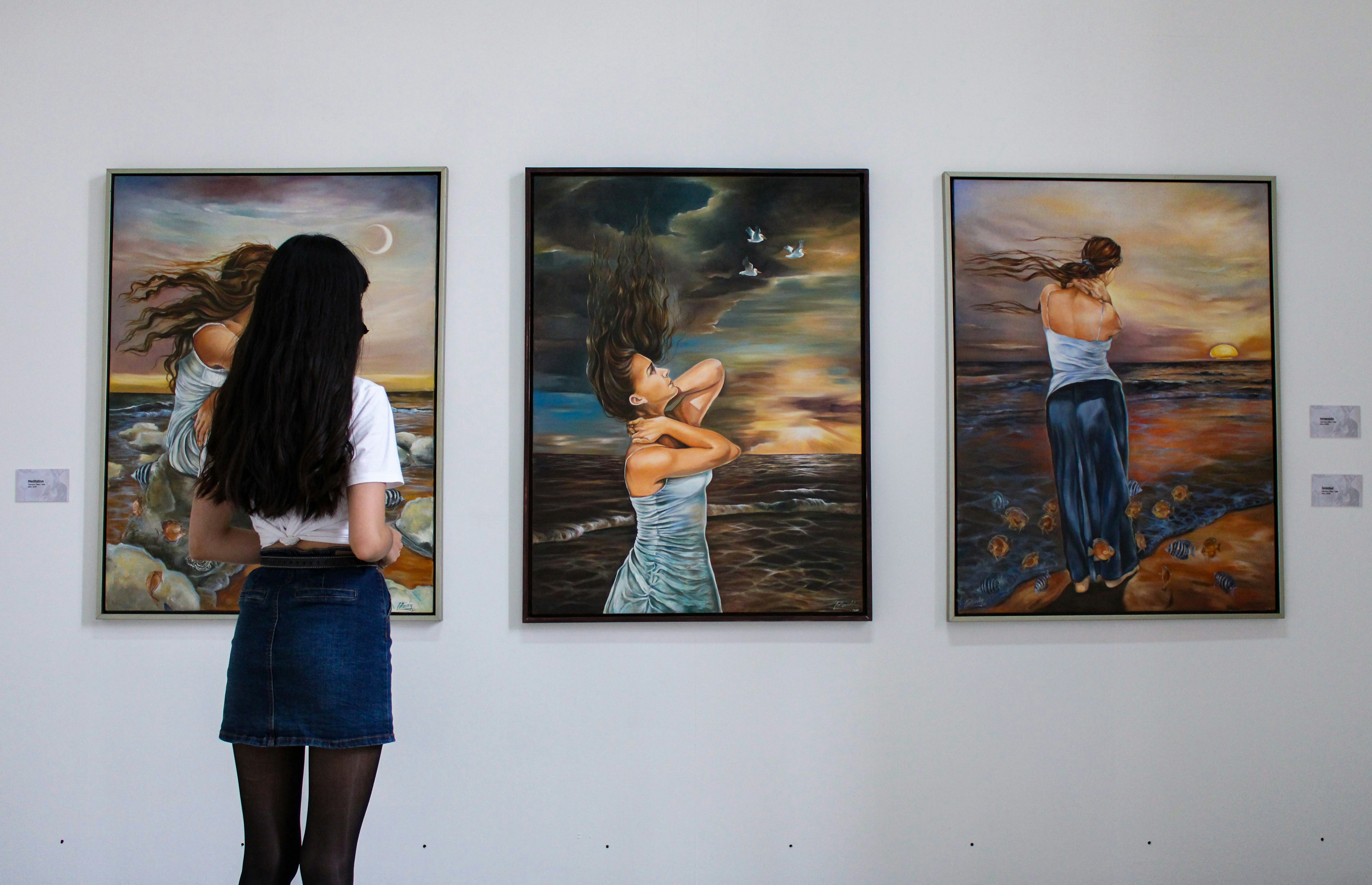 A gallery visitor studies three paintings featuring women against sunset backdrops; the scene captures stillness and contemplation in a minimalist white-walled space.