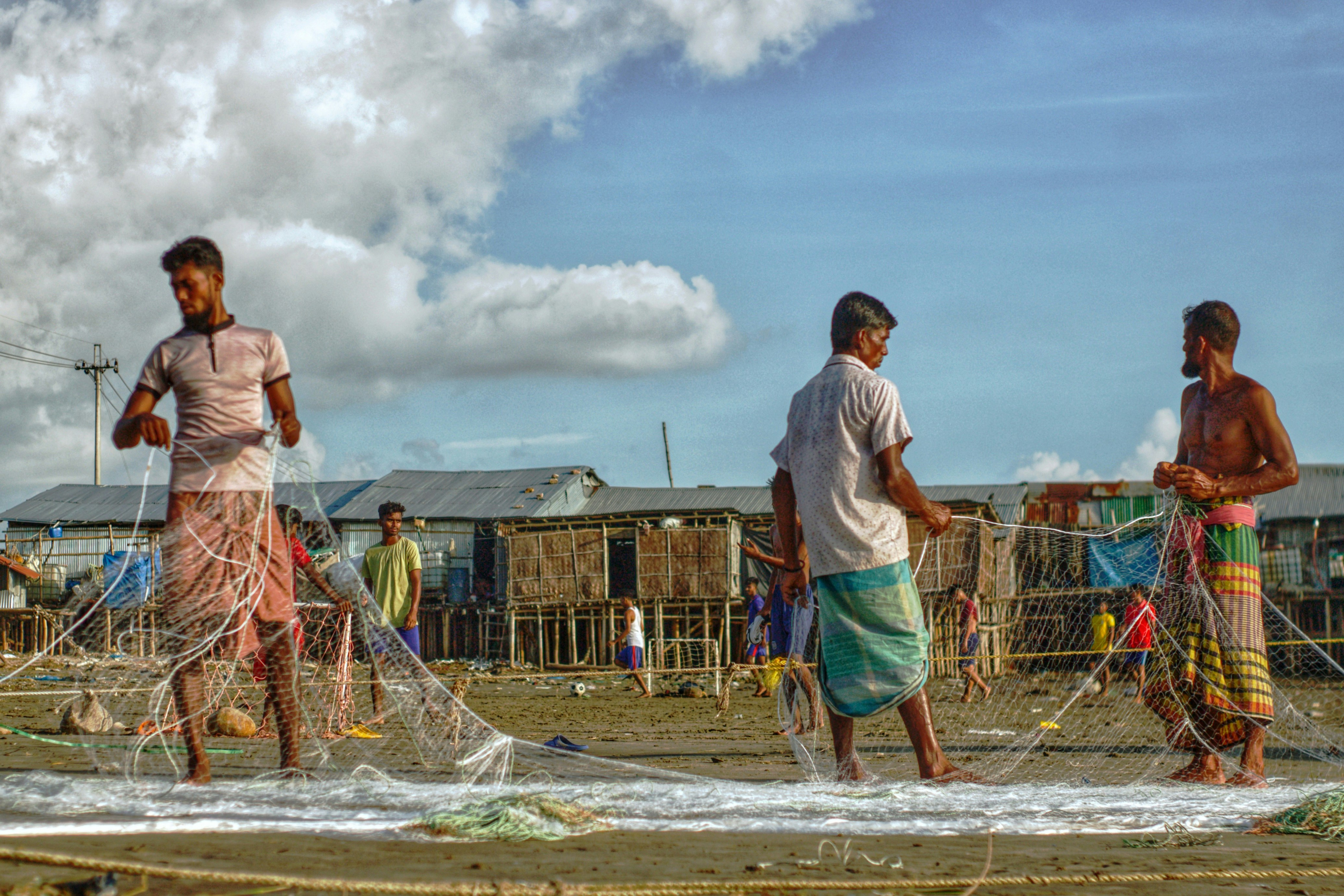Group of men handling fishing nets on a sandy shore with modest huts and a bright sky in the background; a documentary-style scene of coastal livelihoods. (Photograph) 