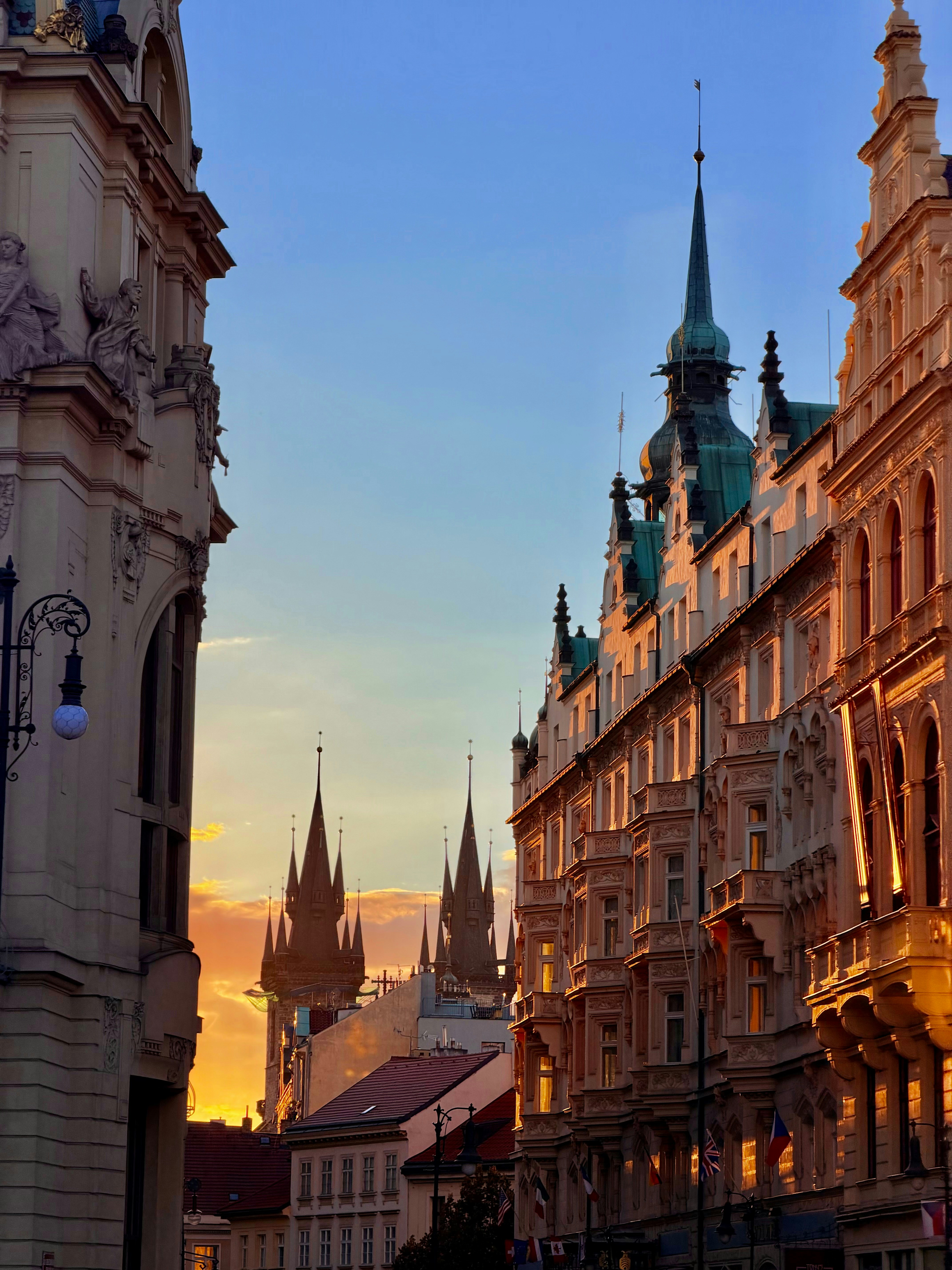 Golden-hour light bathes a European street lined with ornate facades and spires, leading toward a distant church silhouette under a blue-to-orange sky.