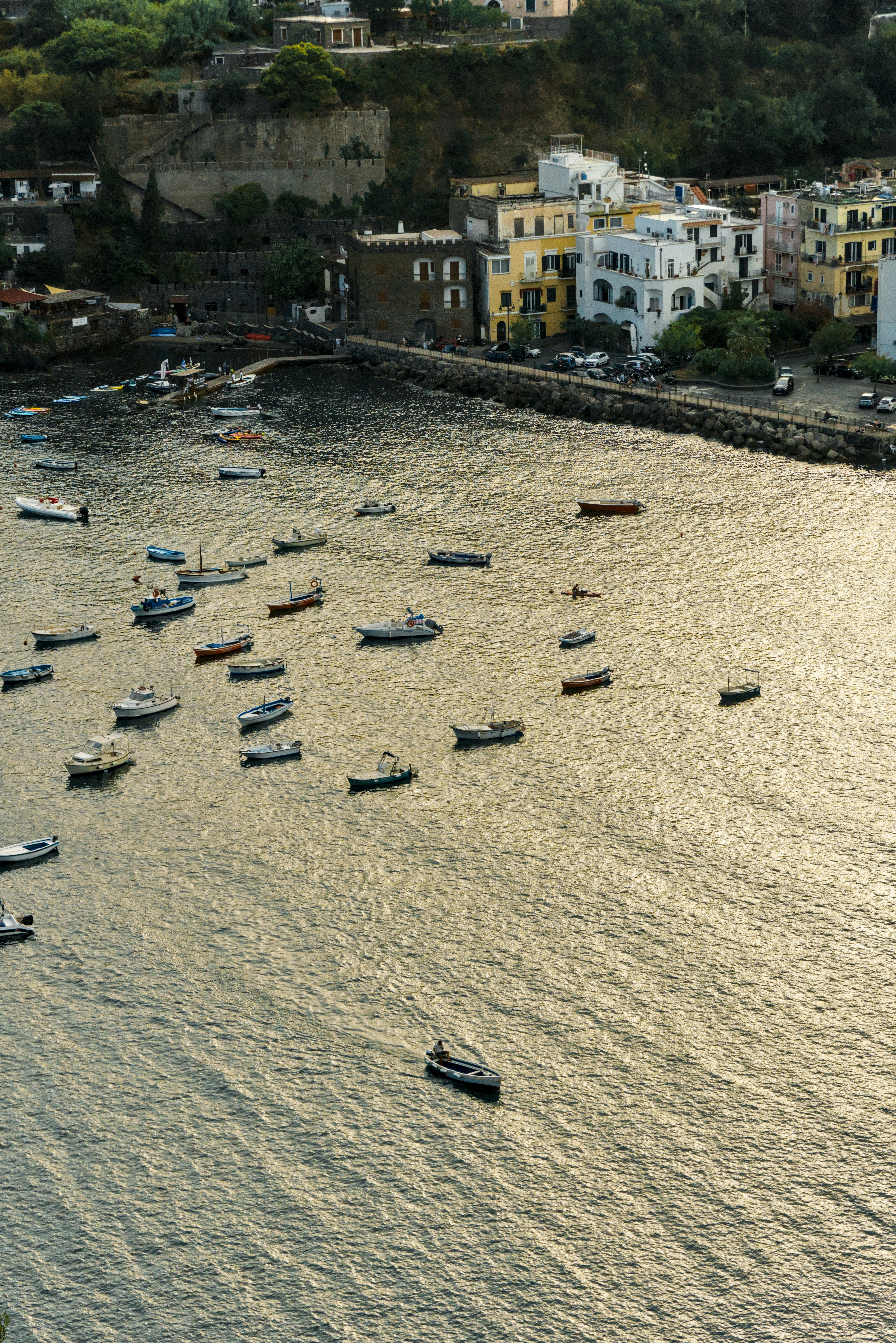 Small boats dot a sunlit harbor with a hillside village in the background, capturing a quiet coastal scene with shimmering water. This is a photographic capture, not a painting or illustration.
