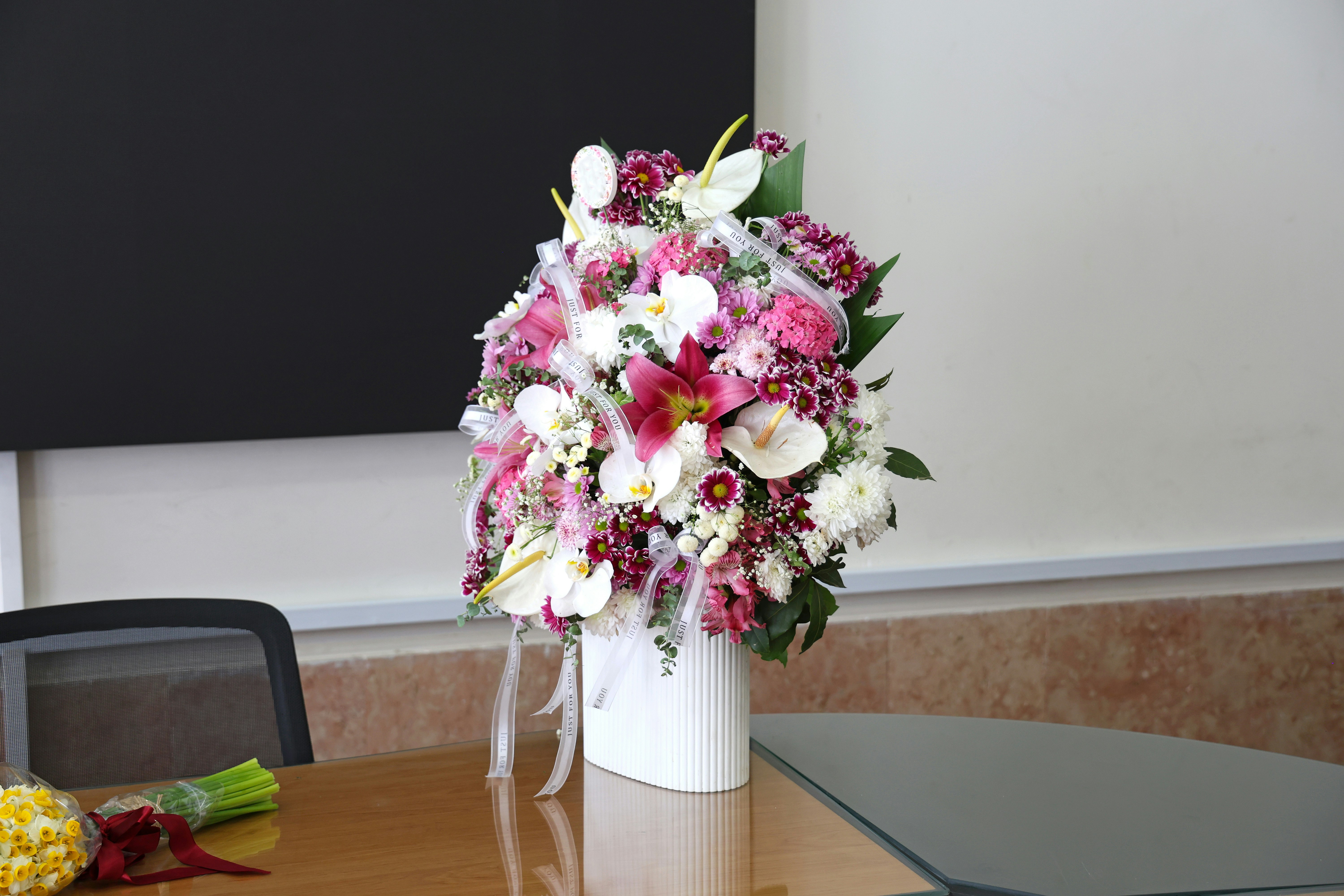 Colorful bouquet in a white vase sits as the focal centerpiece on a wooden table in a room with a black panel and light wall; one laptop or notebook edge is visible nearby.