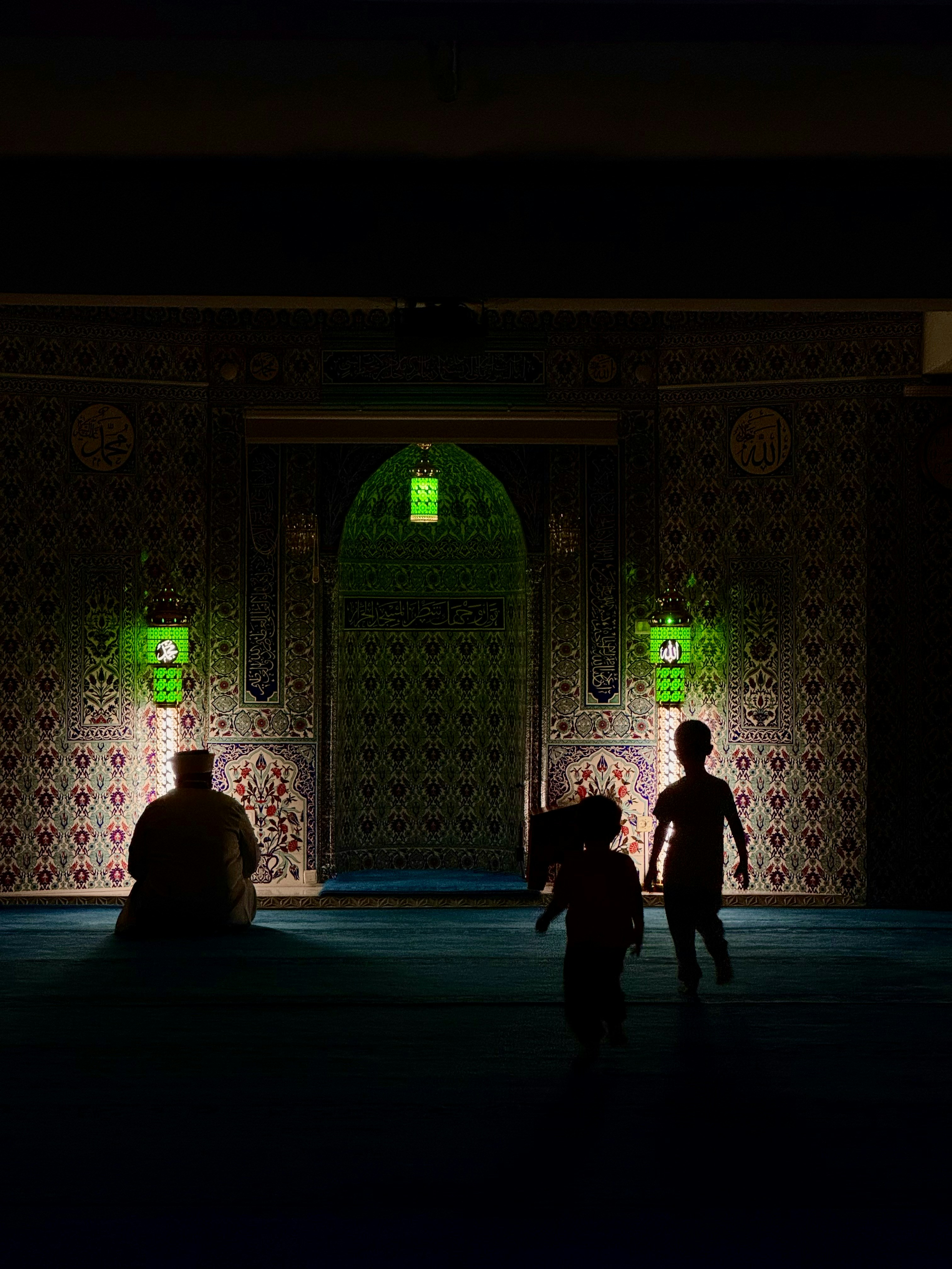 Dimly lit mosque interior with tiled walls and a doorway glowing green; silhouetted figures of a seated person and two children approach the arch, conveying quiet movement inside a sacred space.