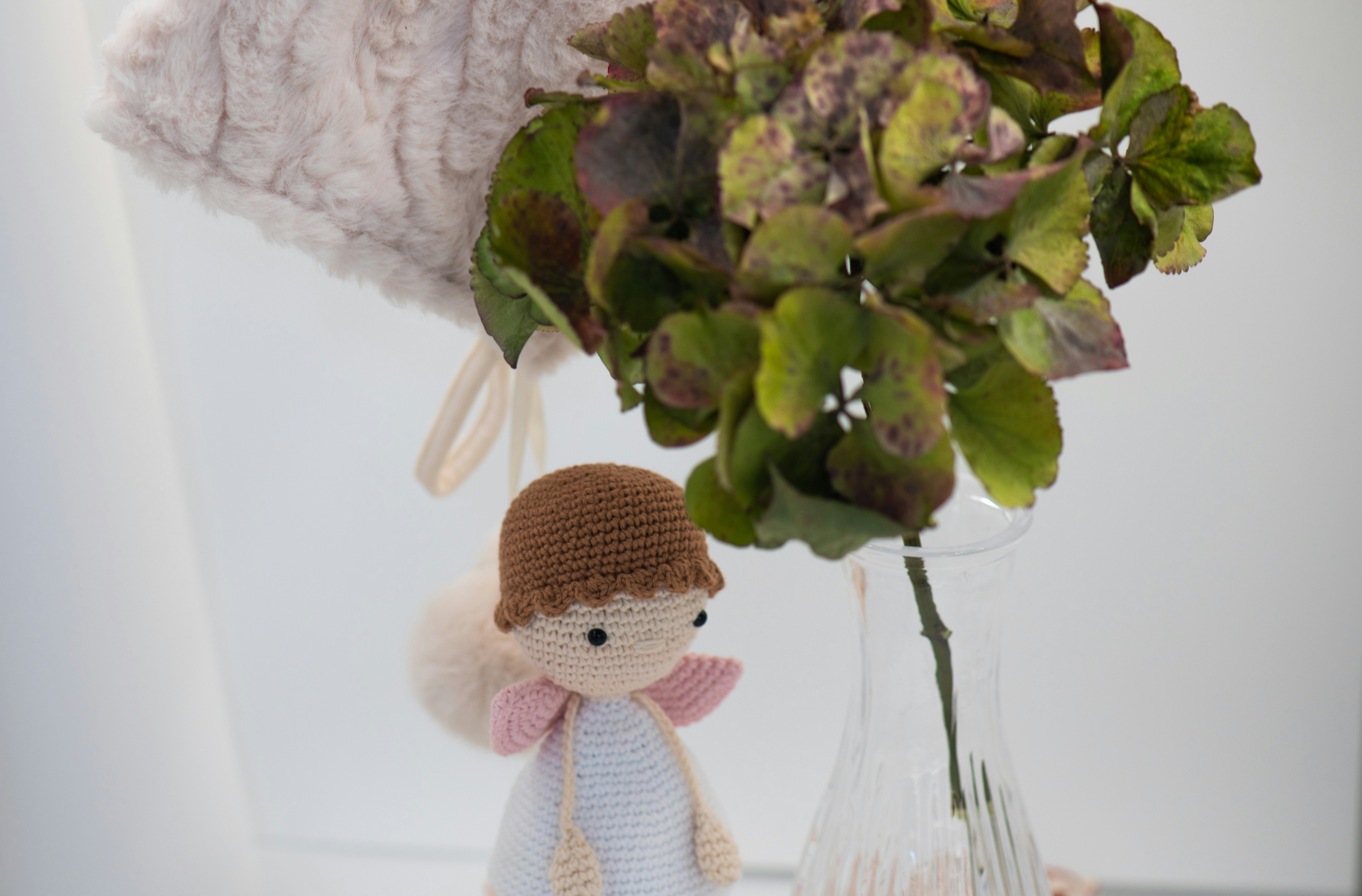 Soft-focus knitted angel and faded hydrangea in a glass vase create a gentle still-life vignette.