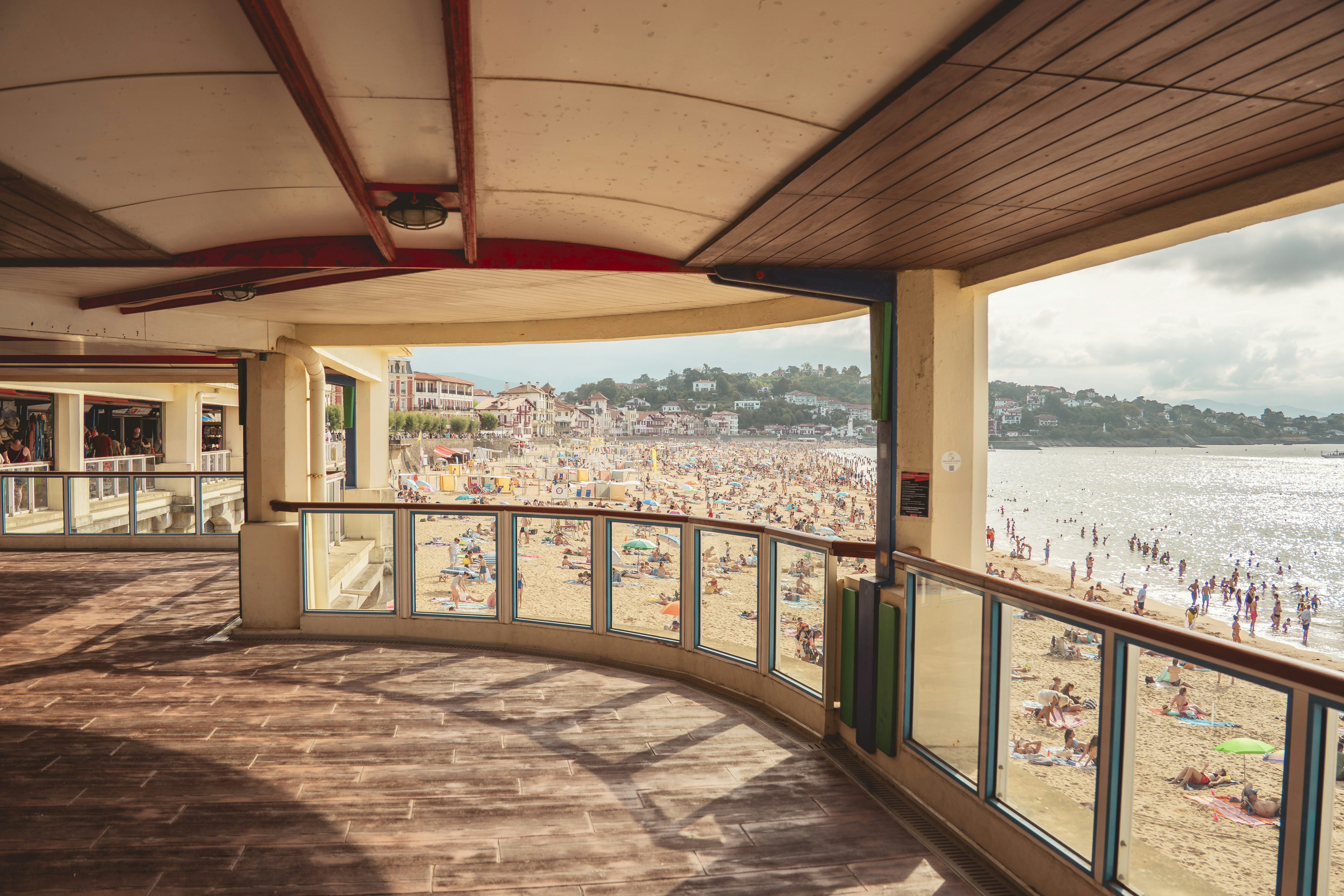 Curved balcony overlooking a crowded beach, warm wooden floor, glass railing, with people and the sea beyond.