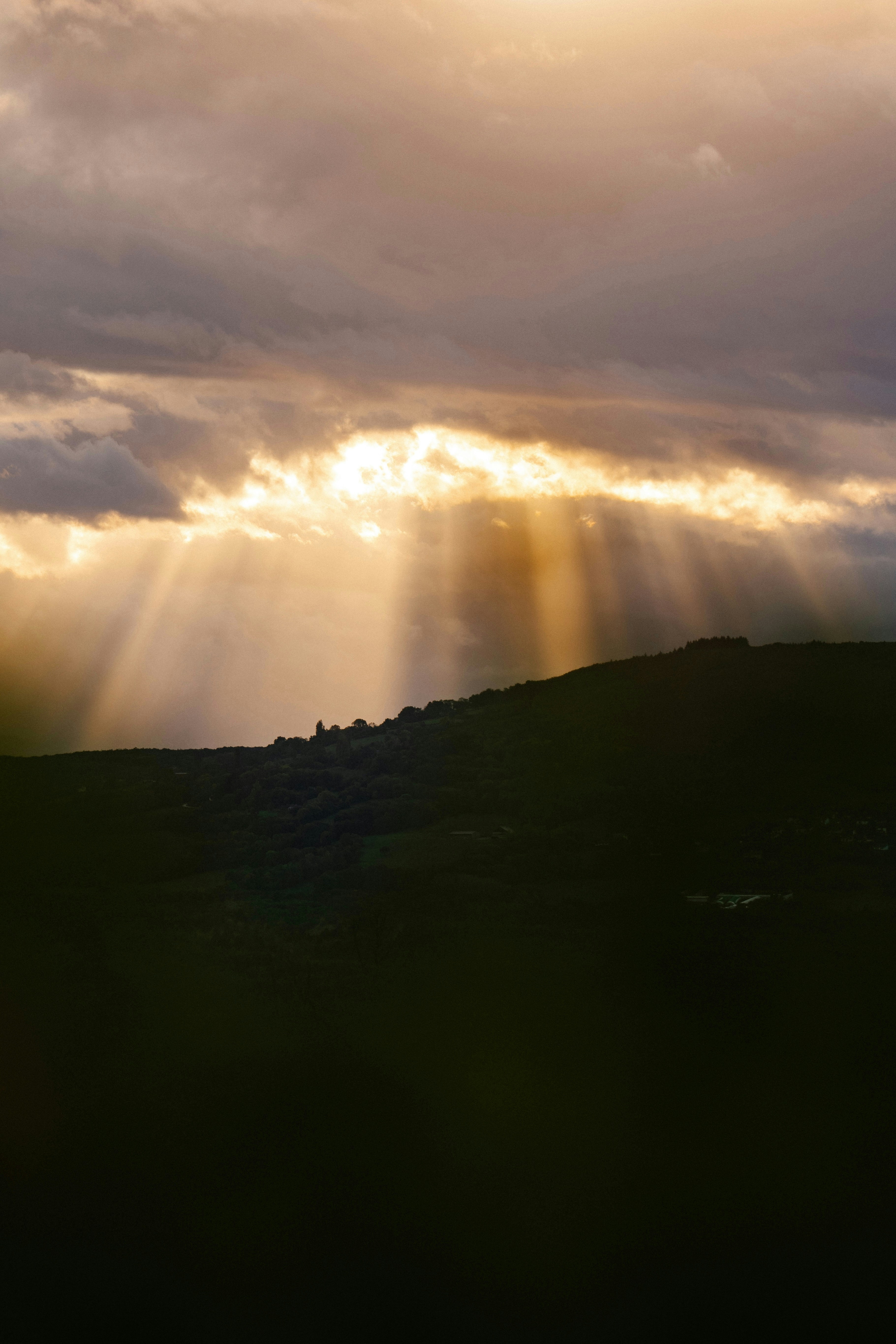 Sunlight streaming through clouds over a dark hillside during the golden hour