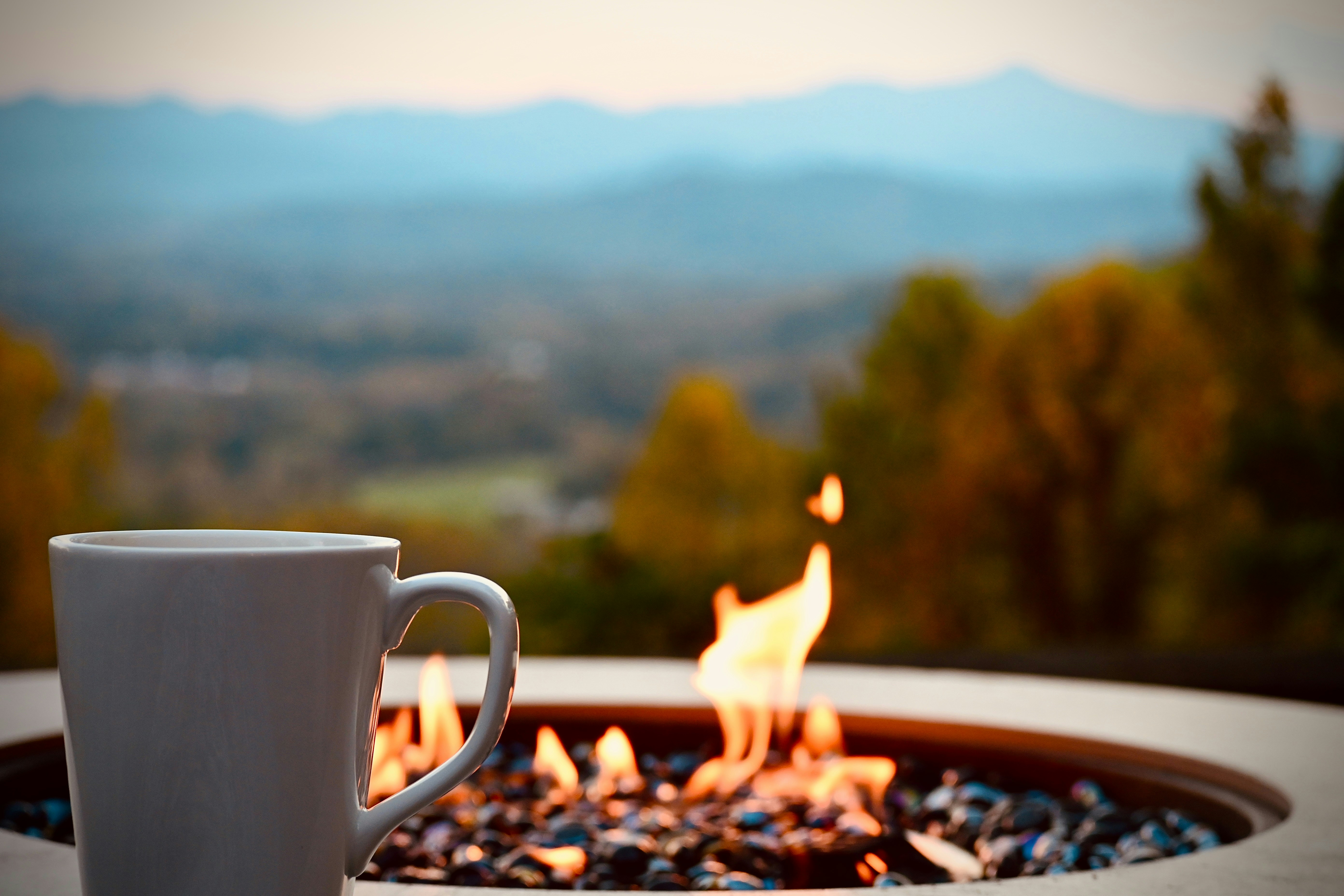 A ceramic mug rests at the edge of a lit fire pit, with a blurred autumn landscape in the background. A cozy moment captured outdoors, including the glow of flames and warm tones in nature.