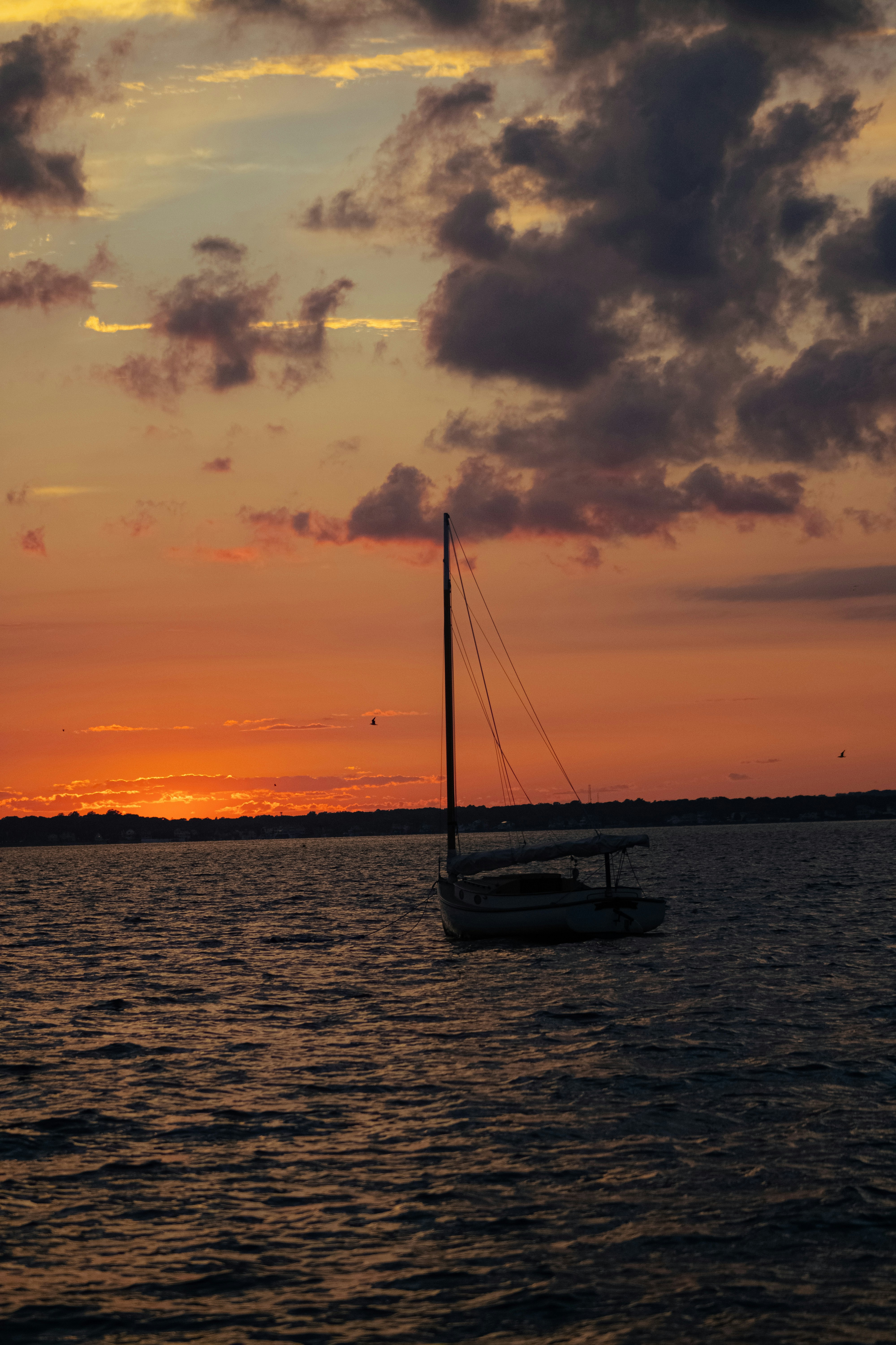 A lone sailboat rests on calm water at sunset, silhouetted against a warm, orange sky. The scene emphasizes tranquil horizon and subtle cloud drama.
