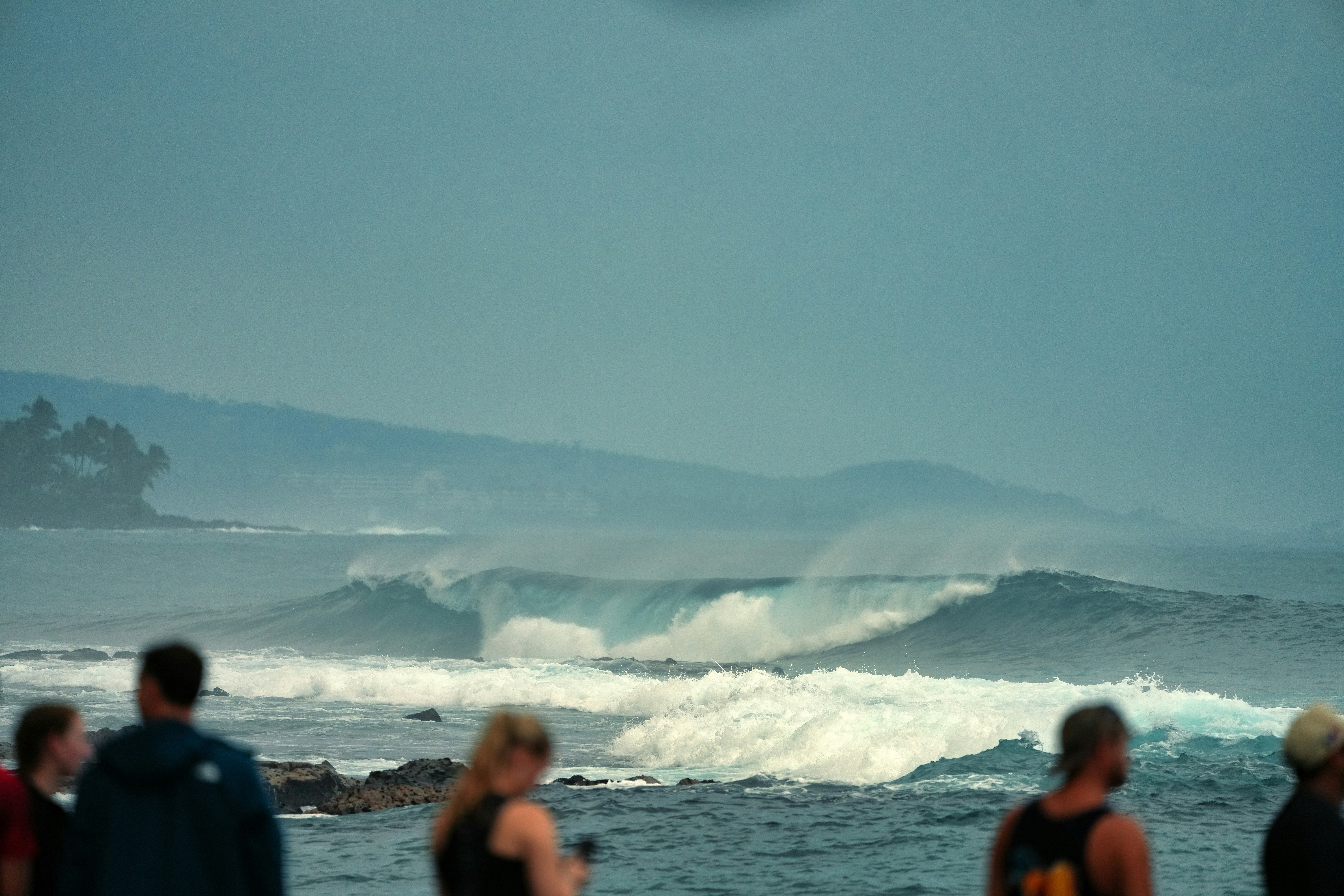 Waves crash near a rocky shoreline with distant hills, while blurred silhouettes of beachgoers fill the foreground.
