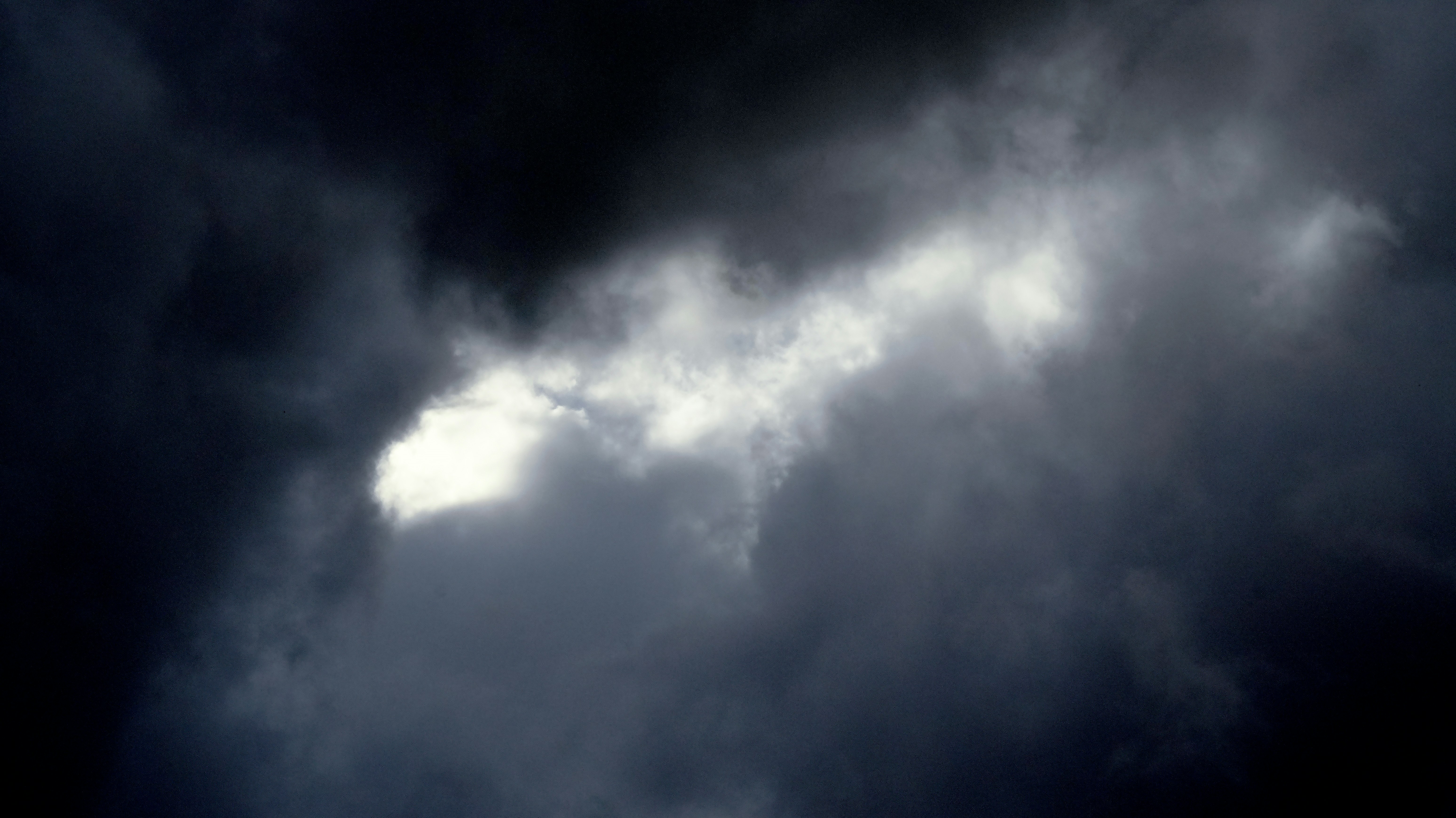 Dark storm clouds part to reveal a solitary patch of light, creating a dramatic contrast and emphasis on the sky’s weight and movement. This image captures natural weather drama without human elements.