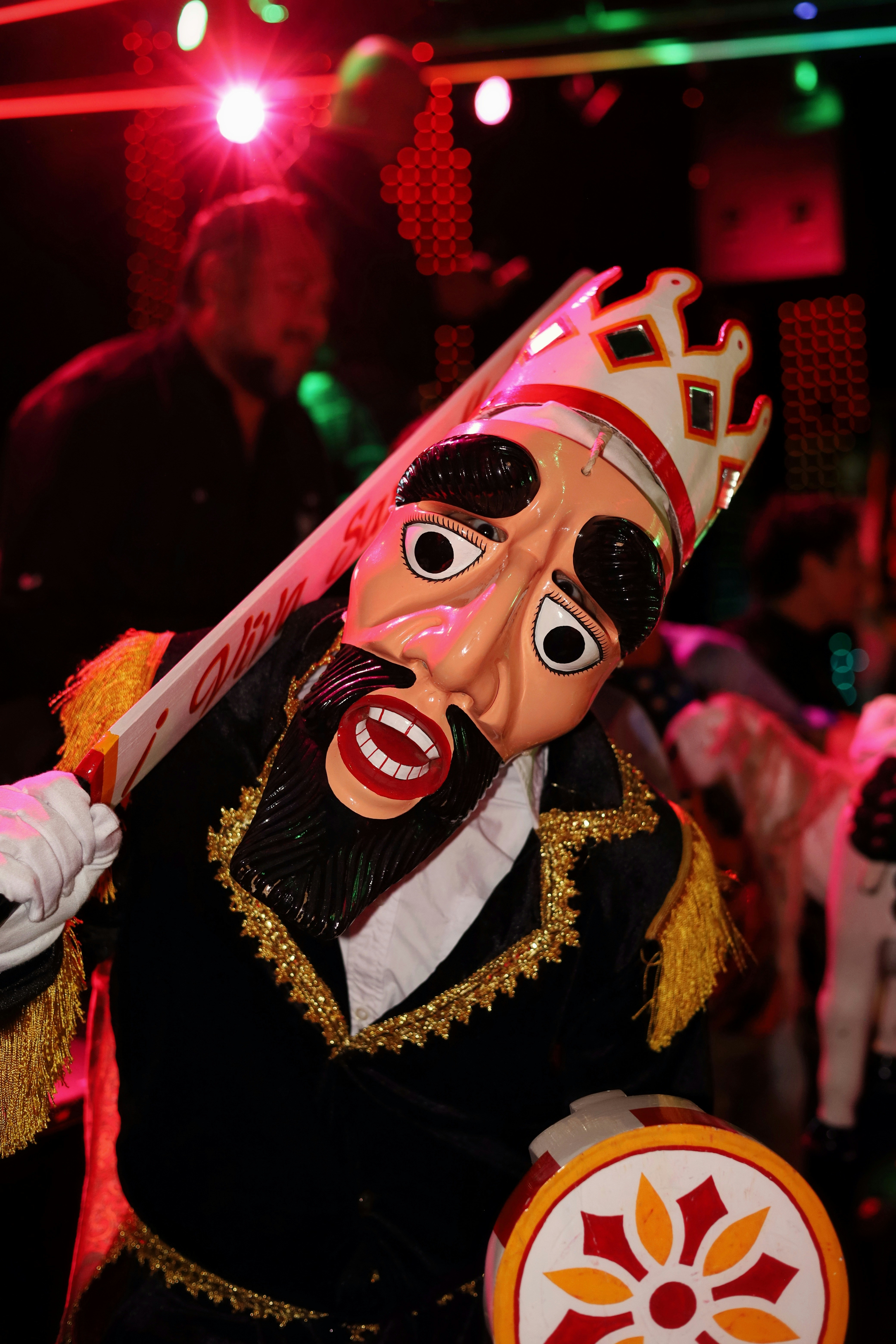Colorful masked performer in a festive costume leads the scene, with a crown mask and bold facial features framed by dynamic lighting.