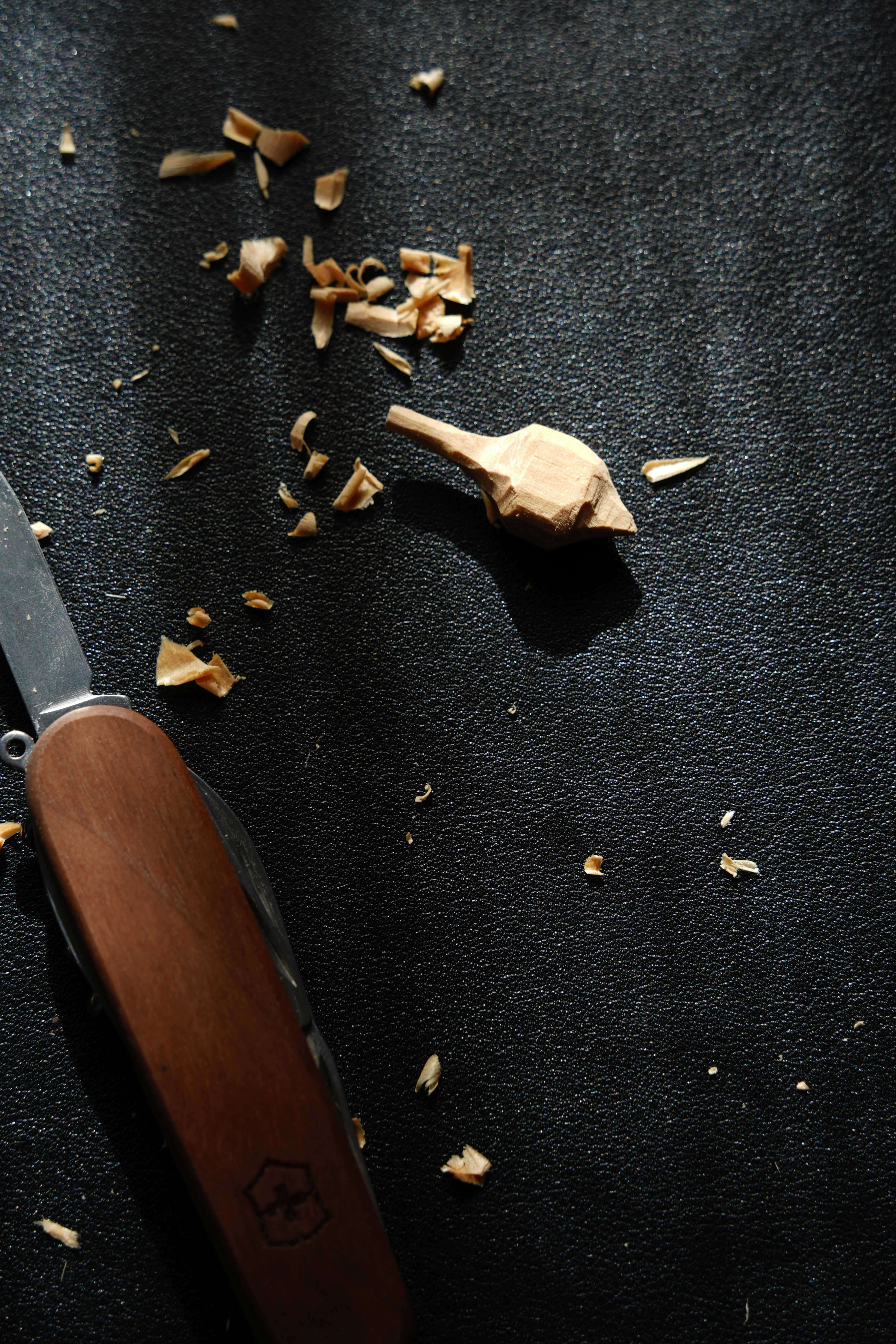A small carved wooden object and scattered wood shavings rest on a dark textured surface, with a portion of a knife handle visible in the lower left. The scene emphasizes manual woodwork and craftsmanship.