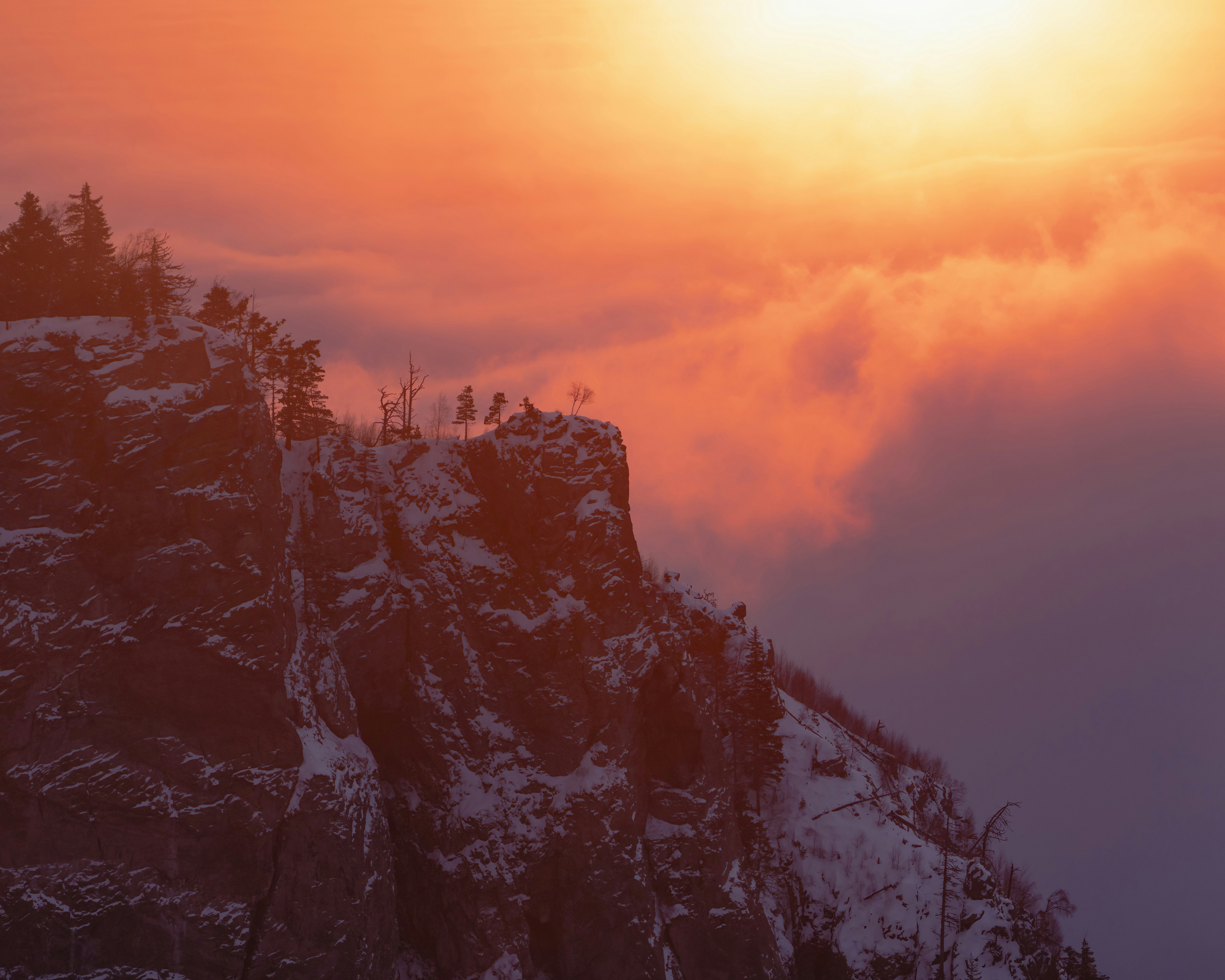 Craggy alpine peak bathed in warm sunset light, half in shadow as orange clouds envelope the horizon. A dramatic landscape with snow-touched rock and a sea of glowing clouds.