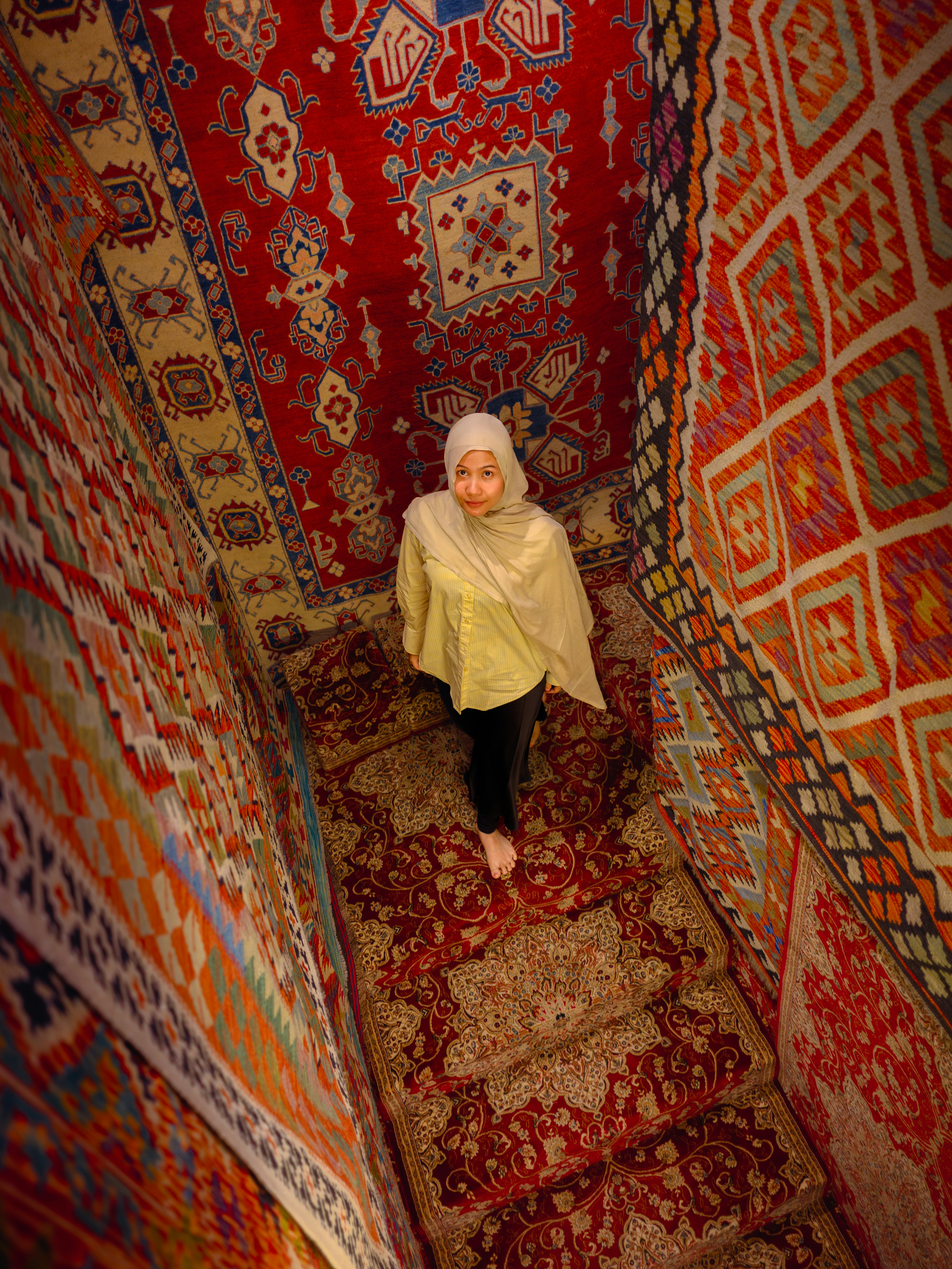 A young woman in a pale hijab stands amid a vividly patterned carpeted stairwell, her presence drawing the eye through the narrow, swirling textile walls.