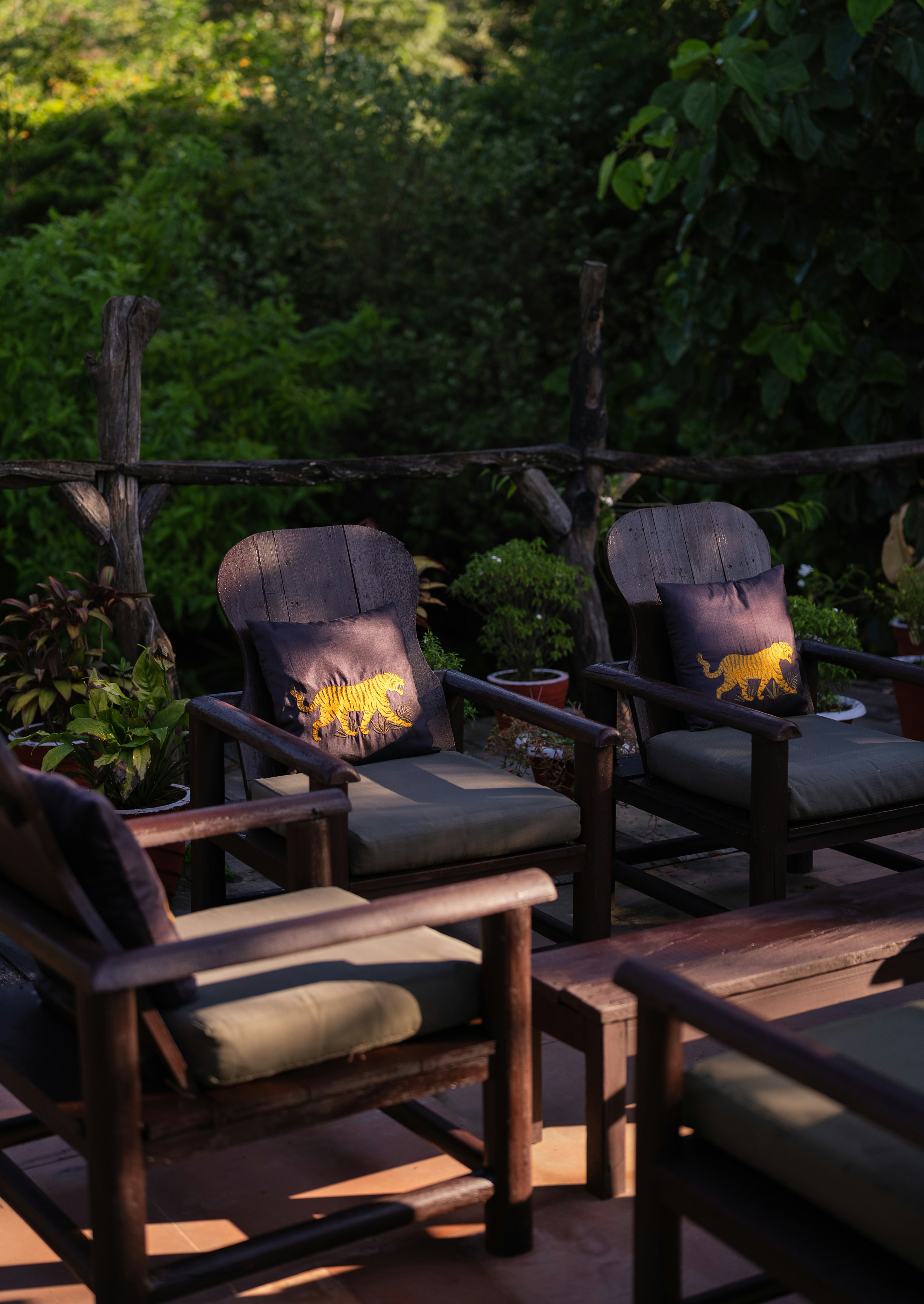 Wooden lounge chairs with dark cushions and yellow animal-embroidered pillows sit in a shaded garden alcove, inviting a tranquil outdoor moment.