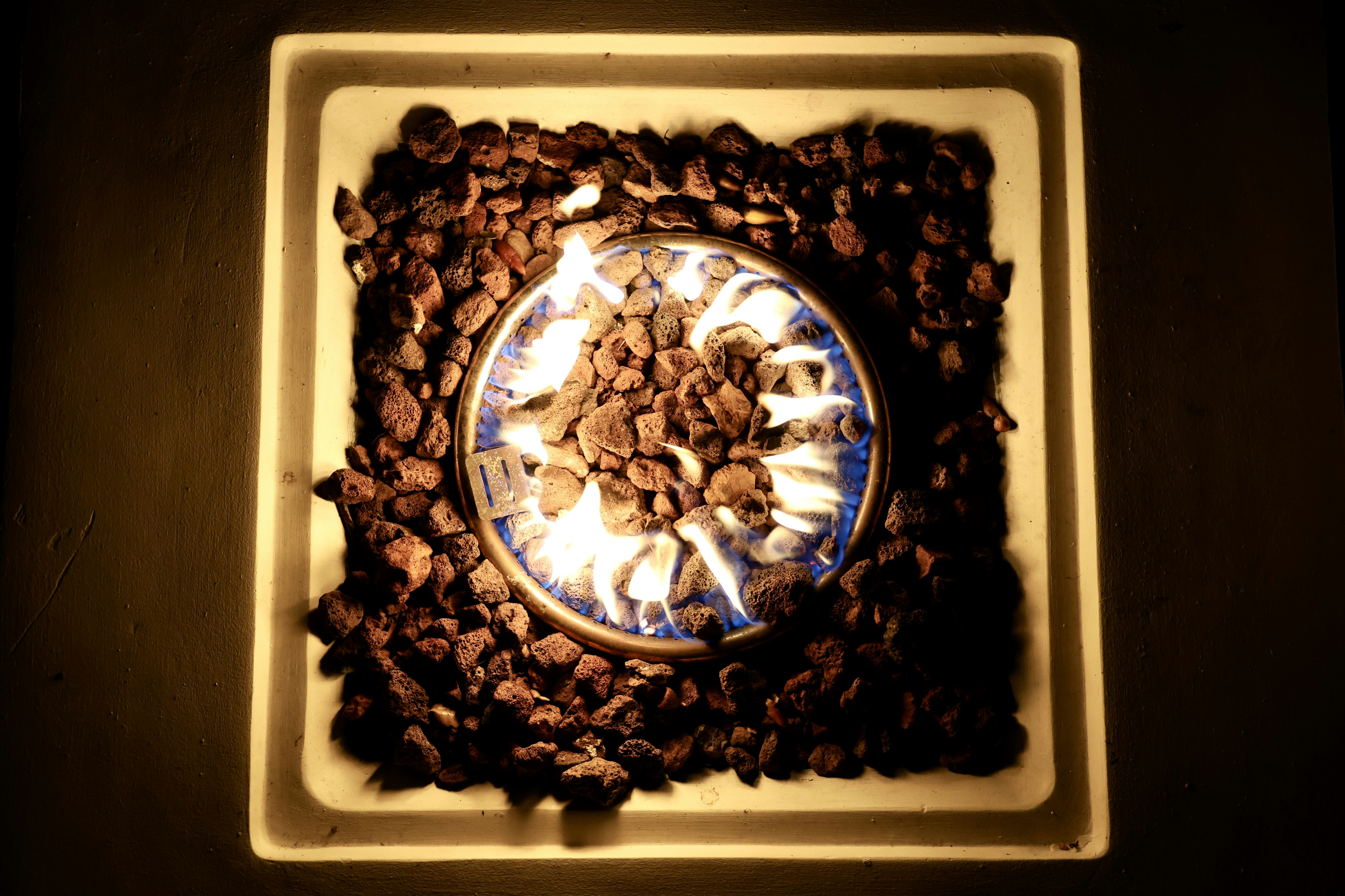 Circular blue burner ring with flames and rocky substrate framed by a square tray; warm lighting highlights texture and depth of the setup.