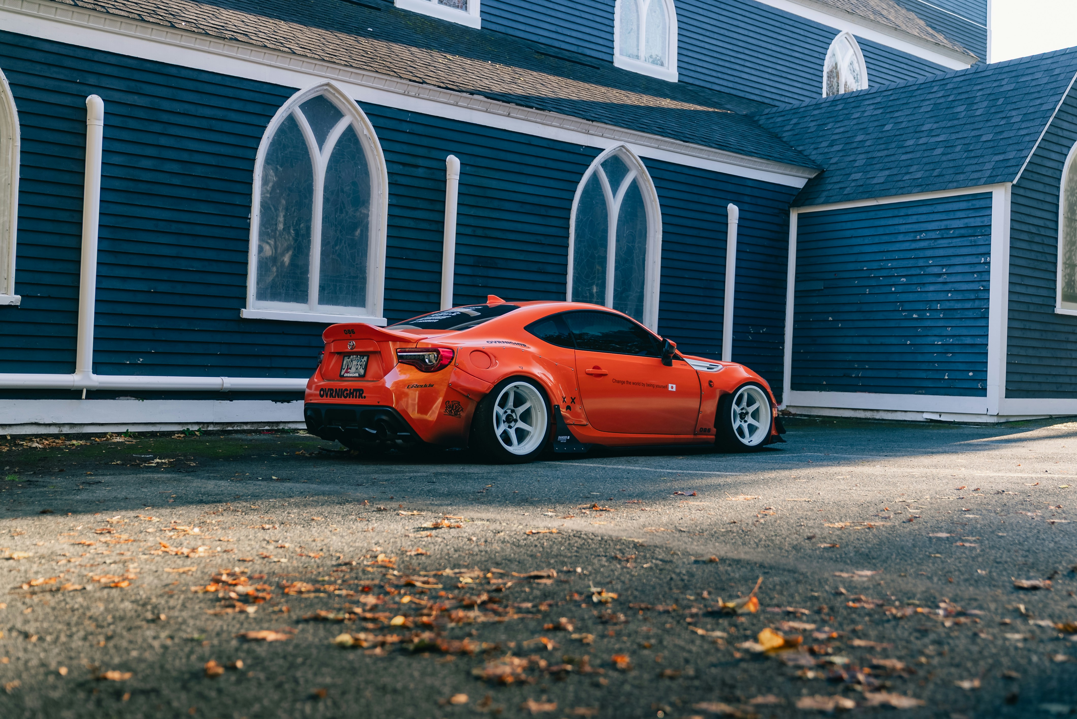 A vivid orange sports car parked beside a blue building with arched windows, grounded by a street and fallen leaves. The scene emphasizes the vehicle as the focal point against a cool architectural backdrop.