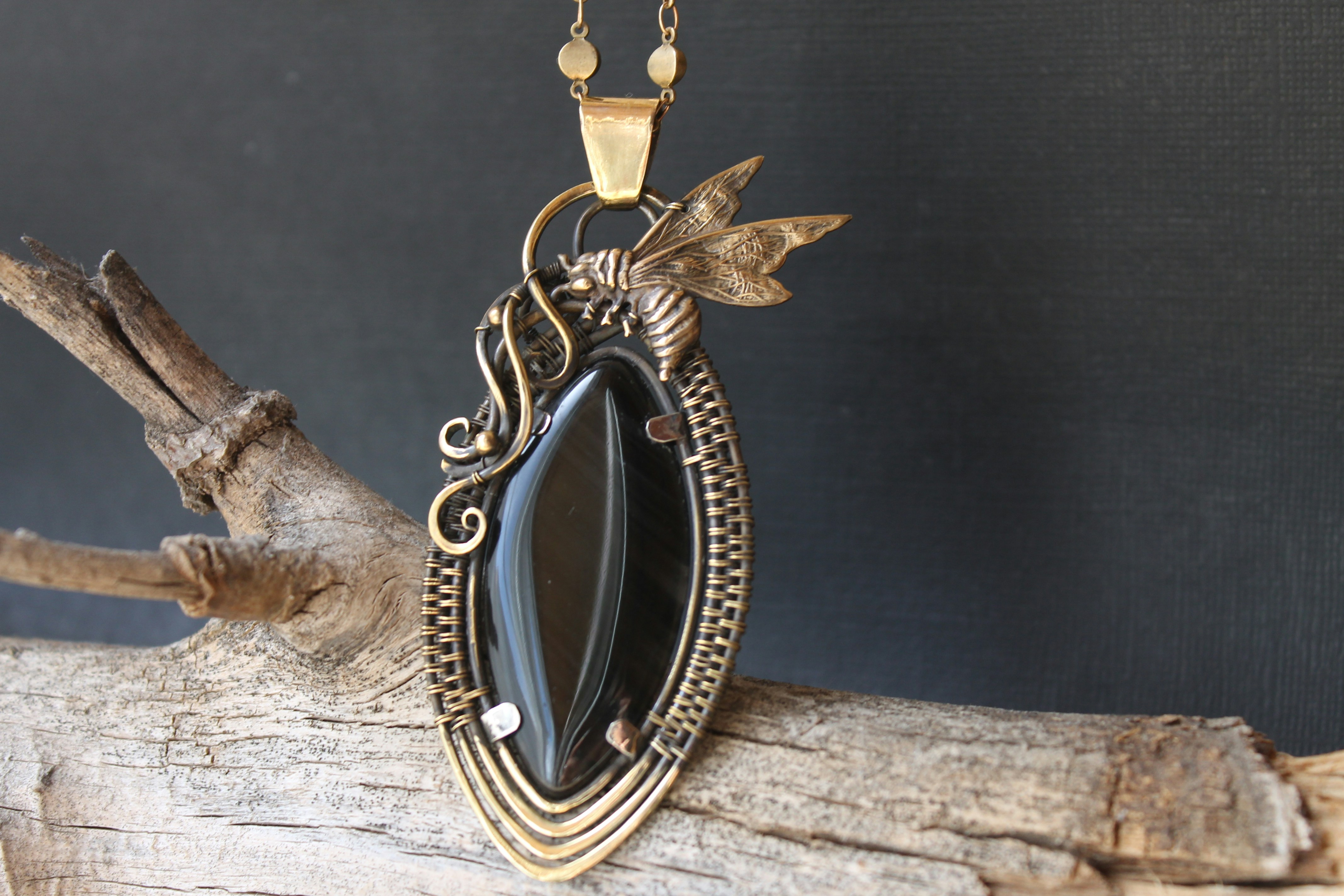 Brass pendant with a dark onyx-like centerpiece hangs from a chain, photographed against a dark backdrop and a rough branch surface.
