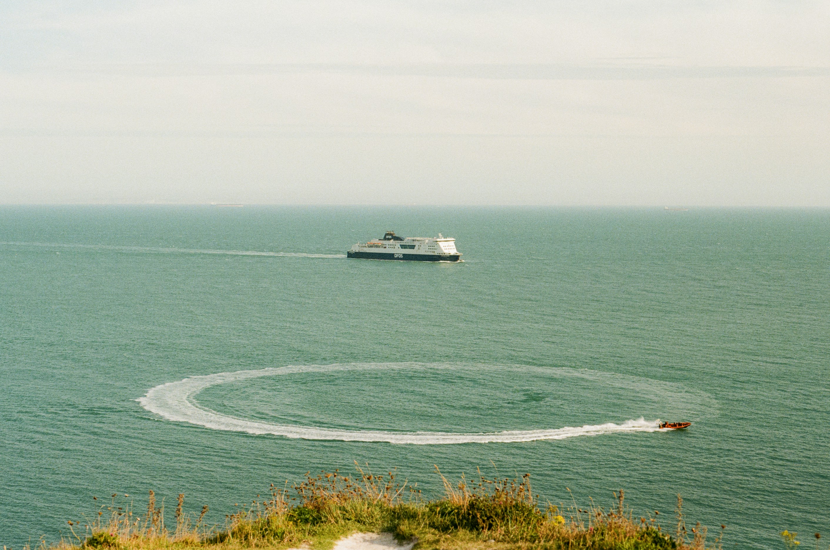 A warm summer day at the White Cliffs of Dover, where the turquoise sea meets a pale, sunlit sky. A ferry drifts across the distant horizon as a speedboat draws a sweeping circle through the water, leaving a bright white trail against the calm blue green sea. Golden grasses catch the afternoon light along the cliff edge, evoking the feeling of a slow coastal escape, salty air and endless summer. Shot on Kodak Gold 200 film in Dover, Kent, England.