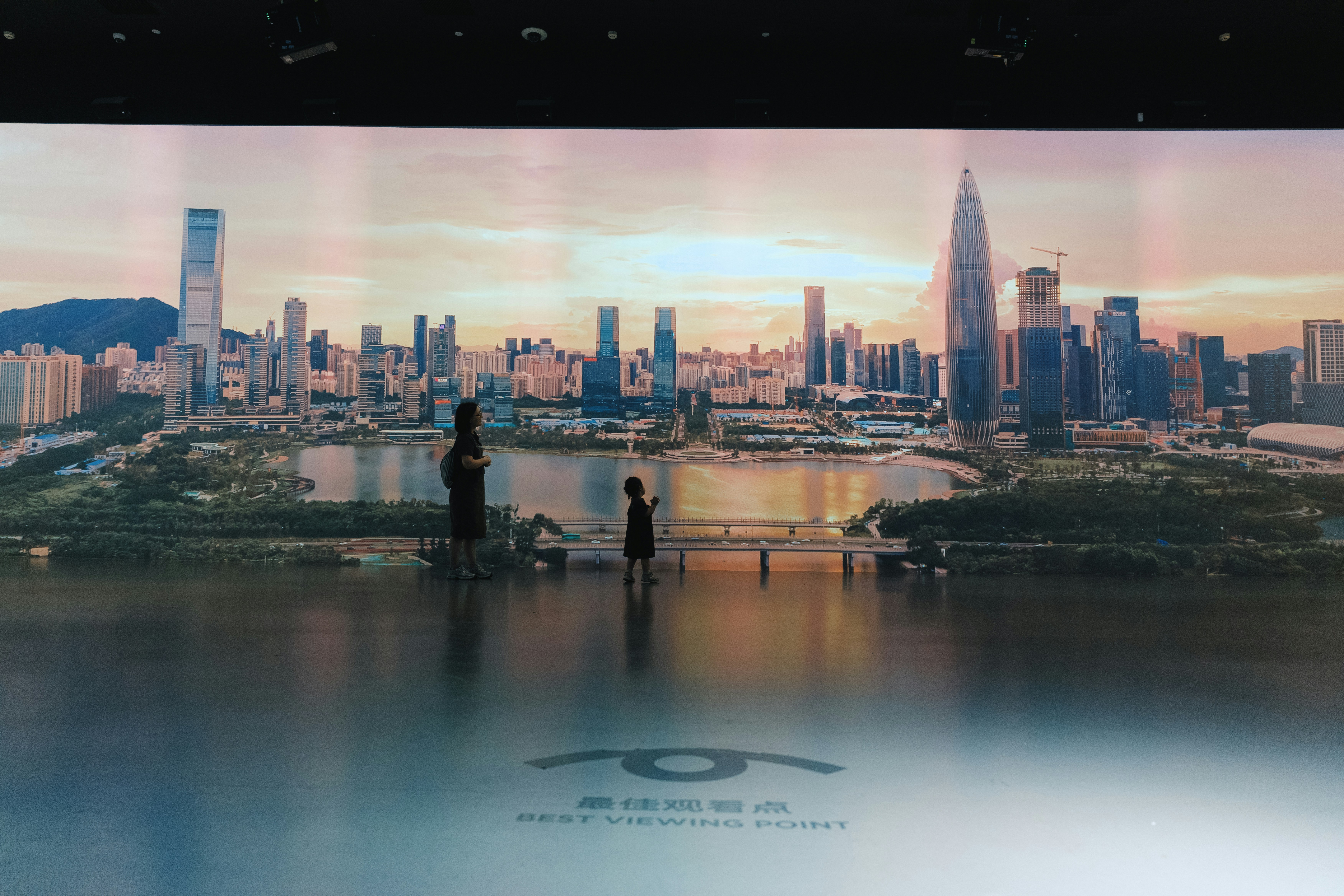 A wide-city panorama with two silhouetted figures standing near a reflective stage backdrop of a modern skyline, highlighting depth and scale in an indoor display environment.