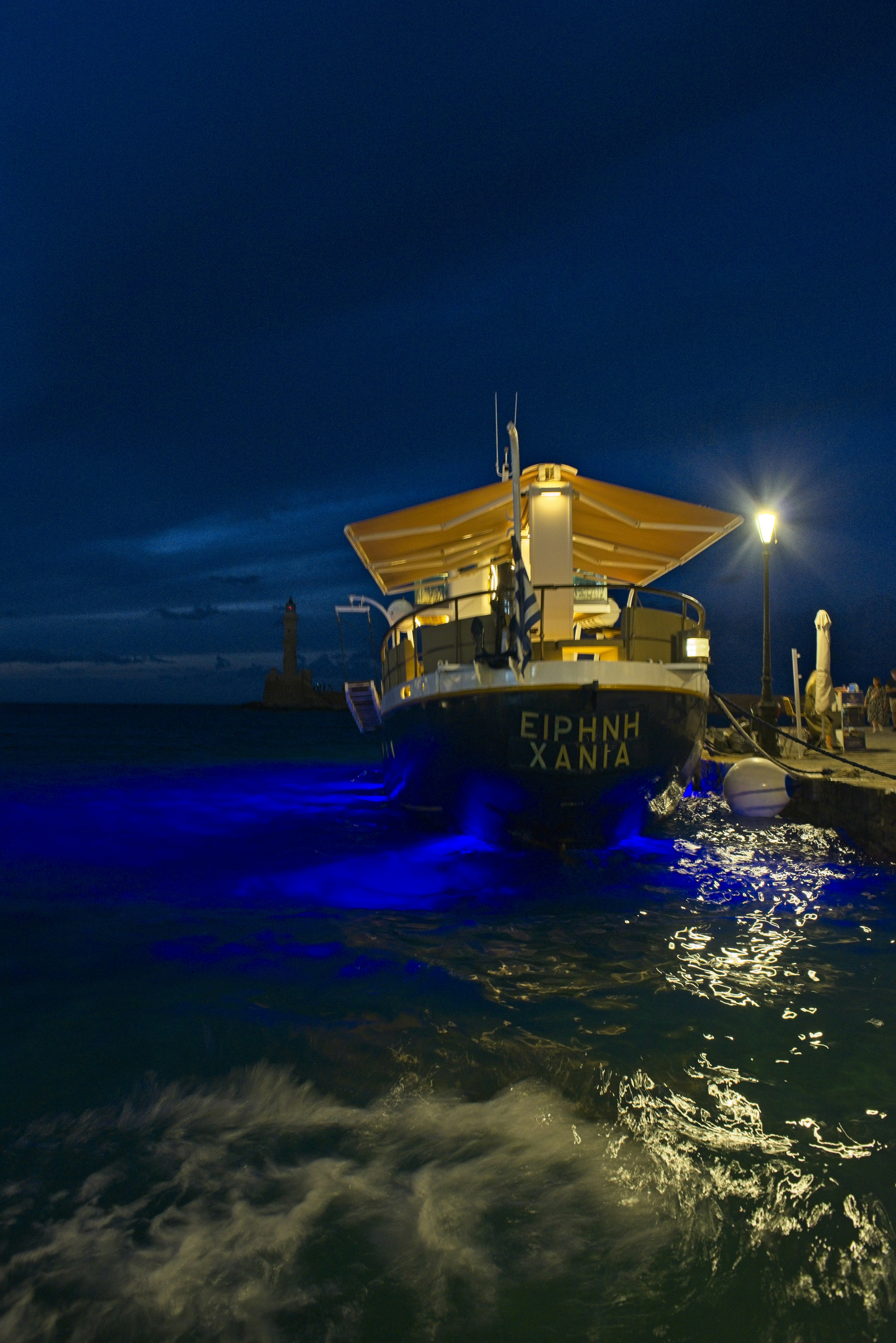 On a windy October evening, the excursion boat “Eirini Chania” rests at the quay of the Old Venetian Harbor, its golden awning glowing beneath a deep indigo sky. Electric-blue underwater lights illuminate the churning water around the hull, while long-exposure waves swirl in the foreground and shimmering reflections dance on the right. A Greek flag flutters at the stern, and a lamp-lit promenade curves beside it. In the distance, the Venetian lighthouse stands as a dark silhouette, completing this vivid, atmospheric blue-hour scene.