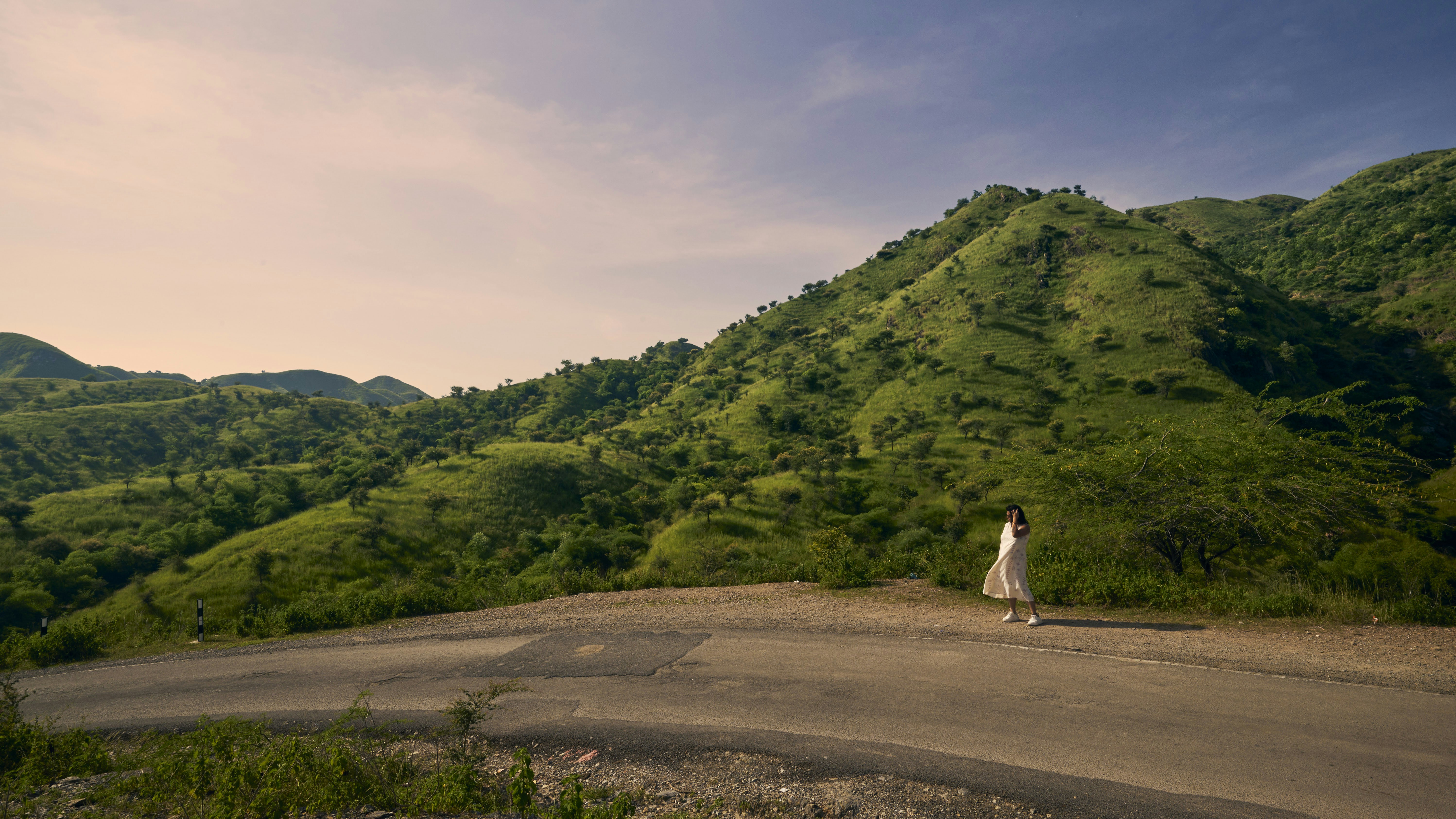 A person in a white dress standing on a winding road through green hills