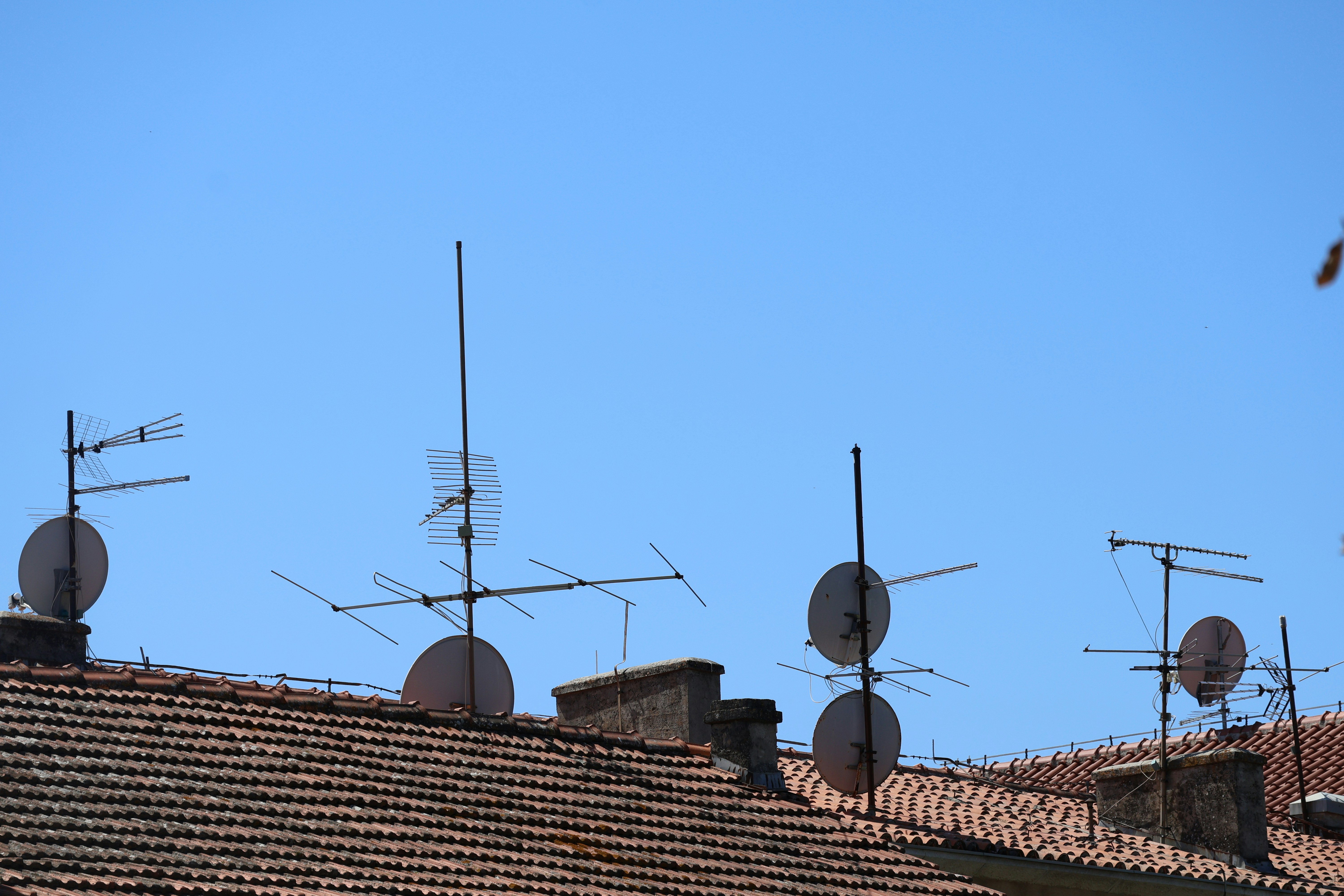 Traditional rooftops of old houses featuring an array of television antennas and satellite dishes clear against a vibrant blue sky. The image captures a classic urban rooftop skyline filled with communication technology and weathered tiles. It brings a nostalgic and rustic aesthetic of telecommunication in daily residential life