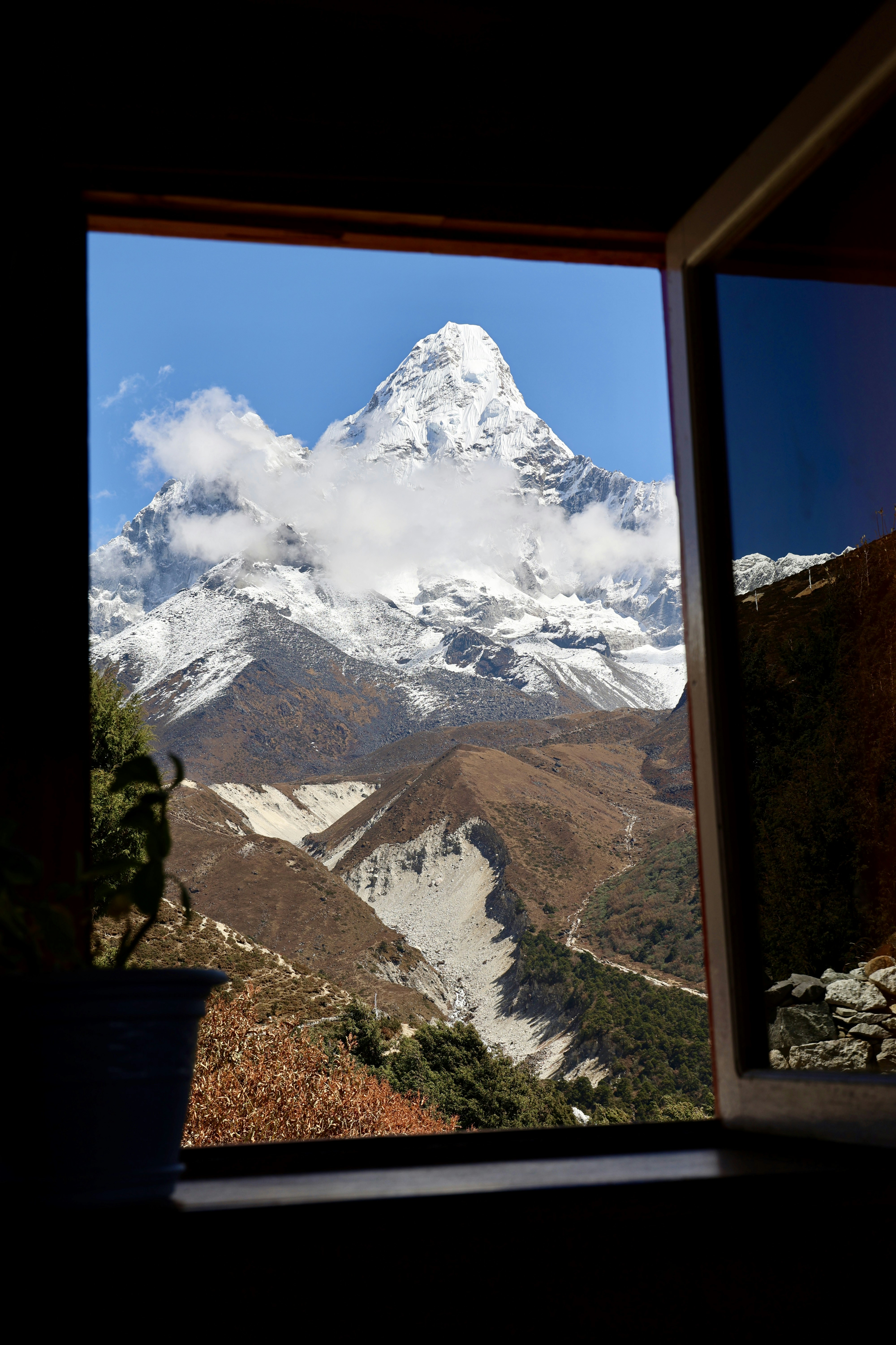 Snow-capped peak framed by an open window with a foreground interior silhouette and a blue sky backdrop. A potted plant sits on the windowsill, highlighting interior contrast with the alpine landscape.