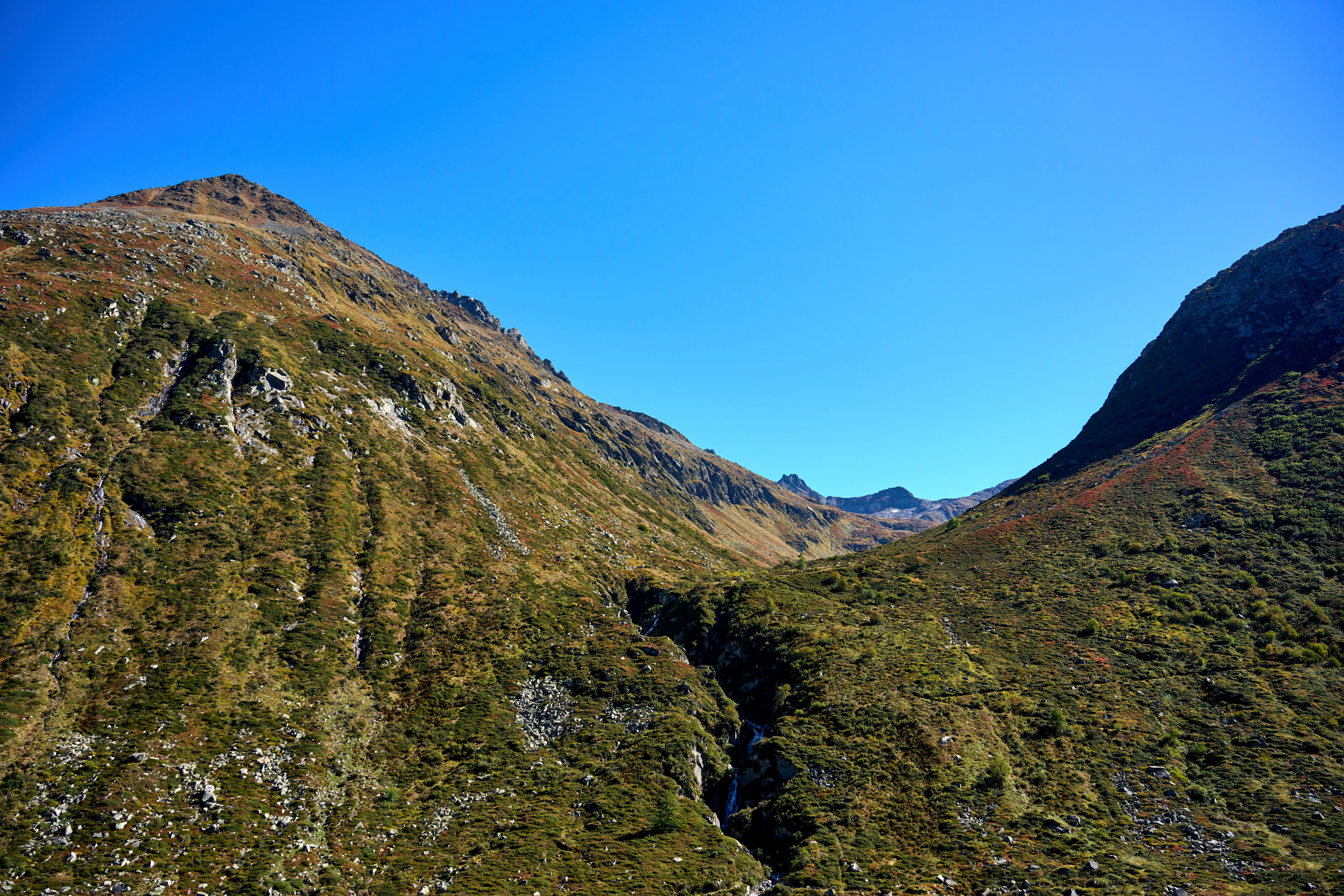 Sweeping mountain valley under a clear blue sky, with rugged slopes and a distant valley corridor. Landscape scene emphasizing textures of moss and rock across a wide, sunlit expanse.